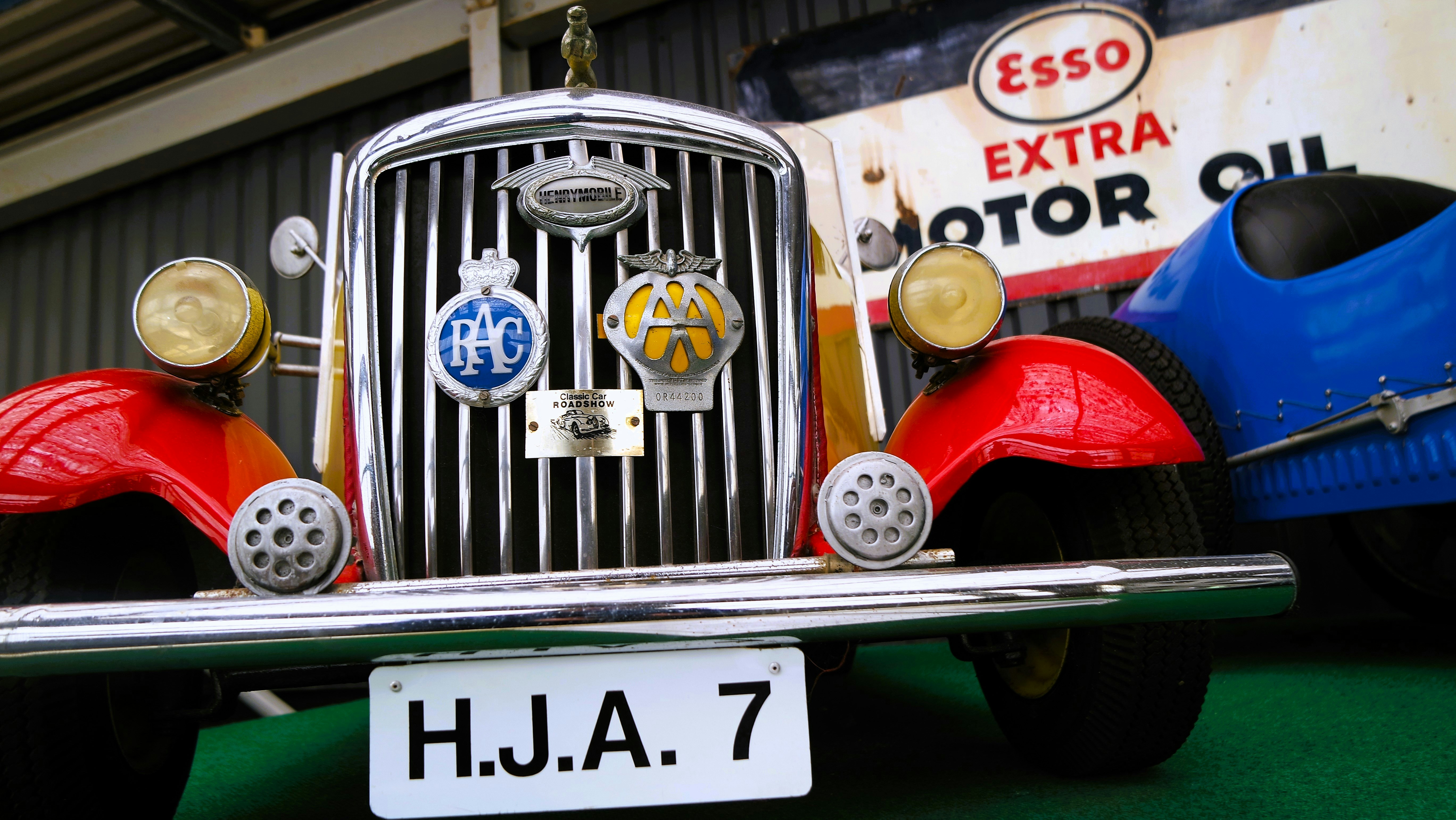 A classic red and chrome car with distinctive badges and a vintage license plate, set against an old Esso motor oil sign. The scene captures the essence of automotive history.
