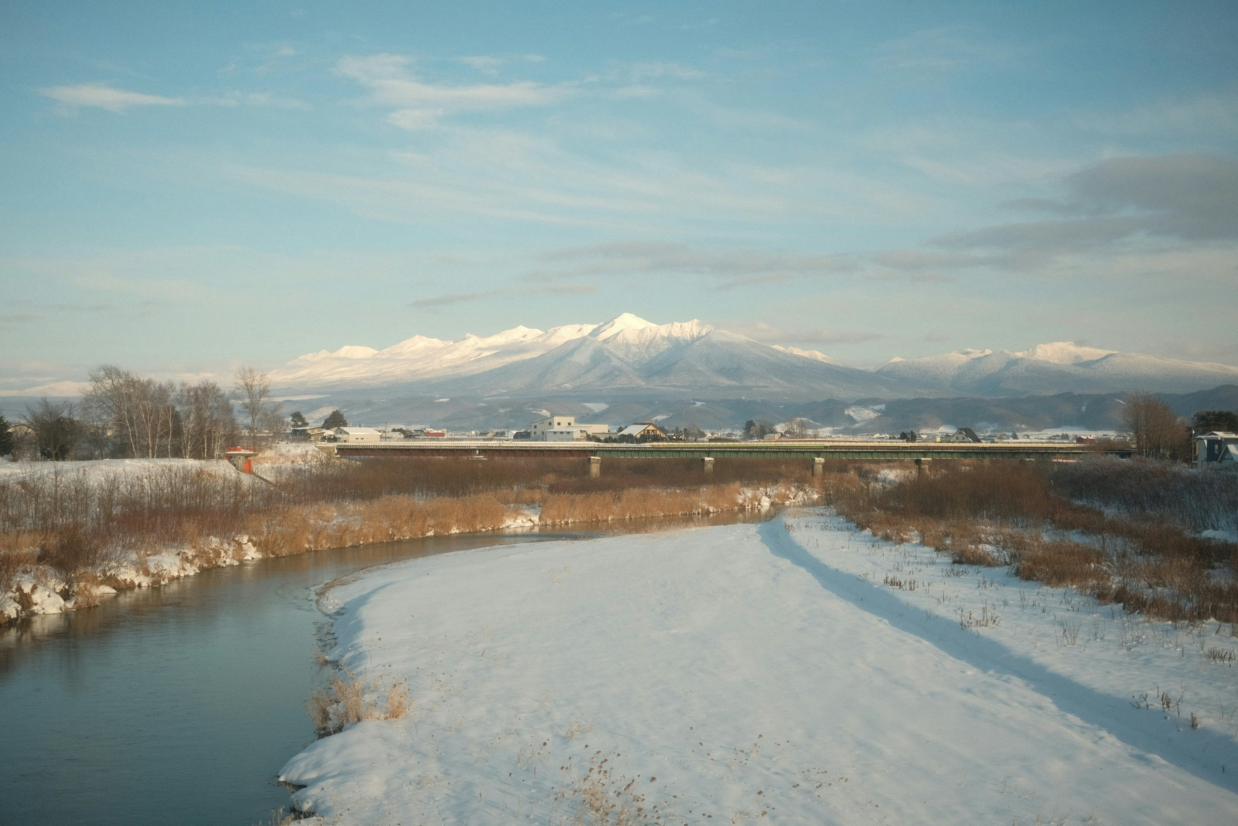 Snow-covered riverbank winding towards distant snow-capped mountains under a soft blue sky.