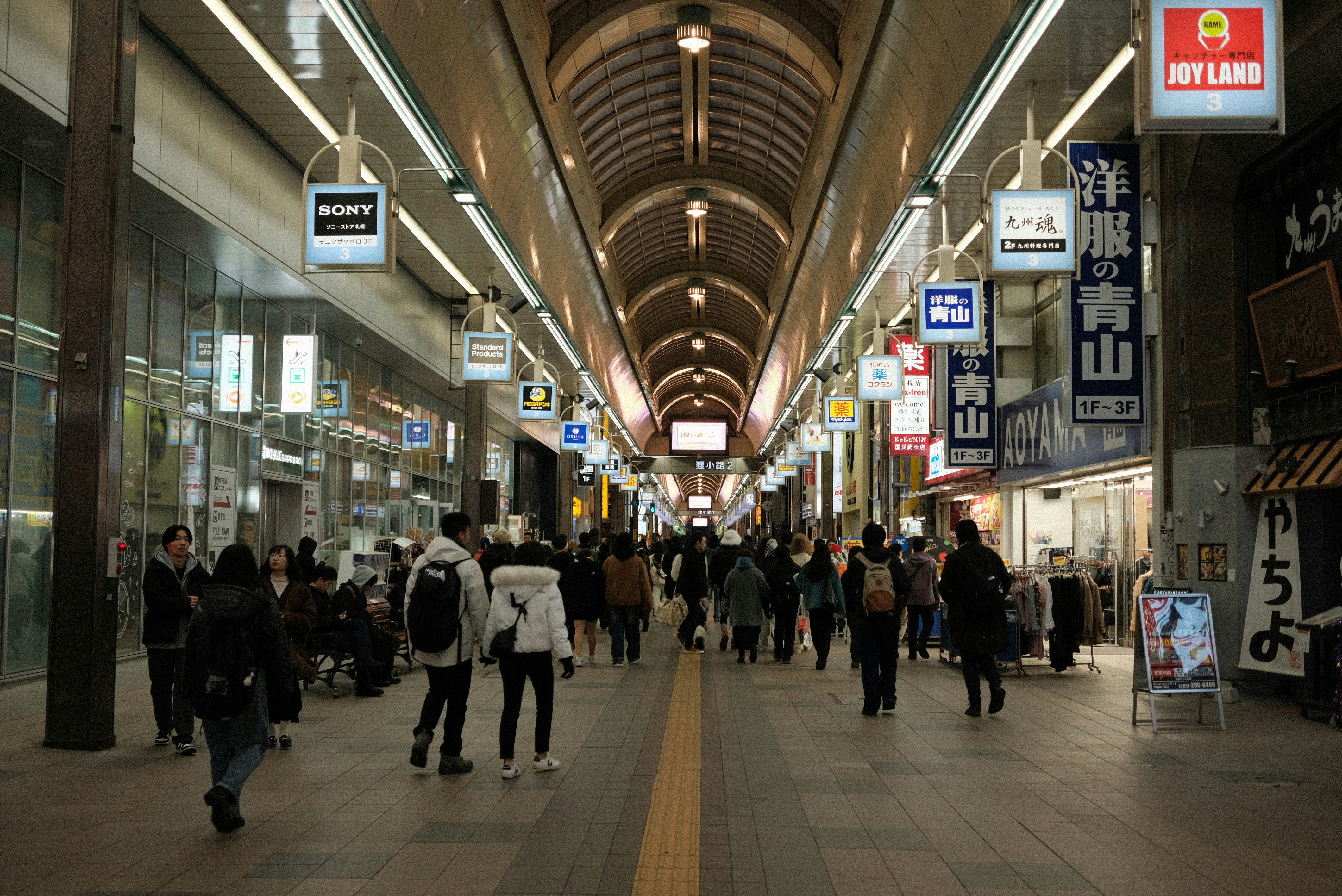 Bustling shopping arcade with people under a warm-toned arched ceiling and colorful illuminated signs.