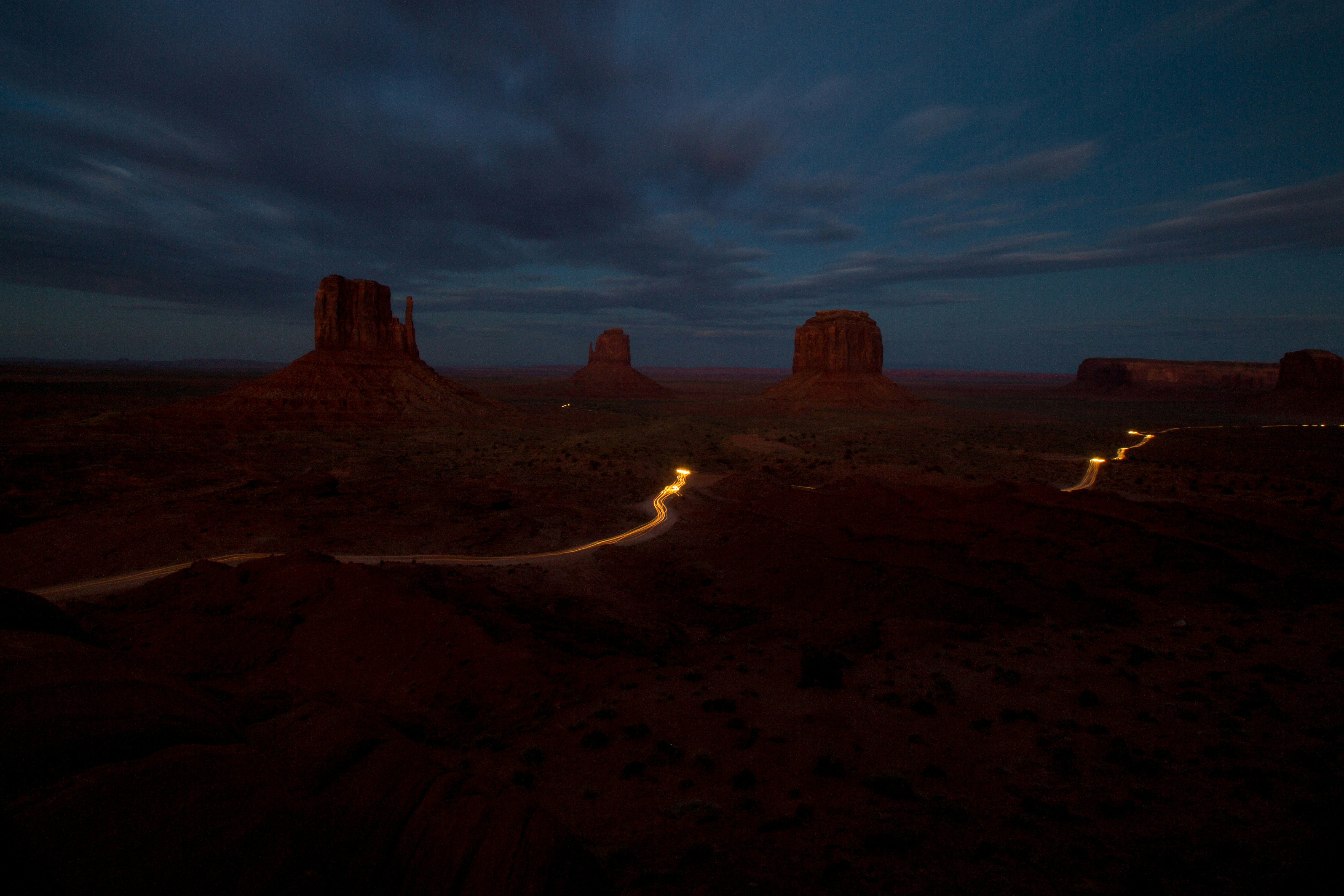 Desert landscape with towering rock formations and a winding trail illuminated by faint lights under a cloudy night sky.