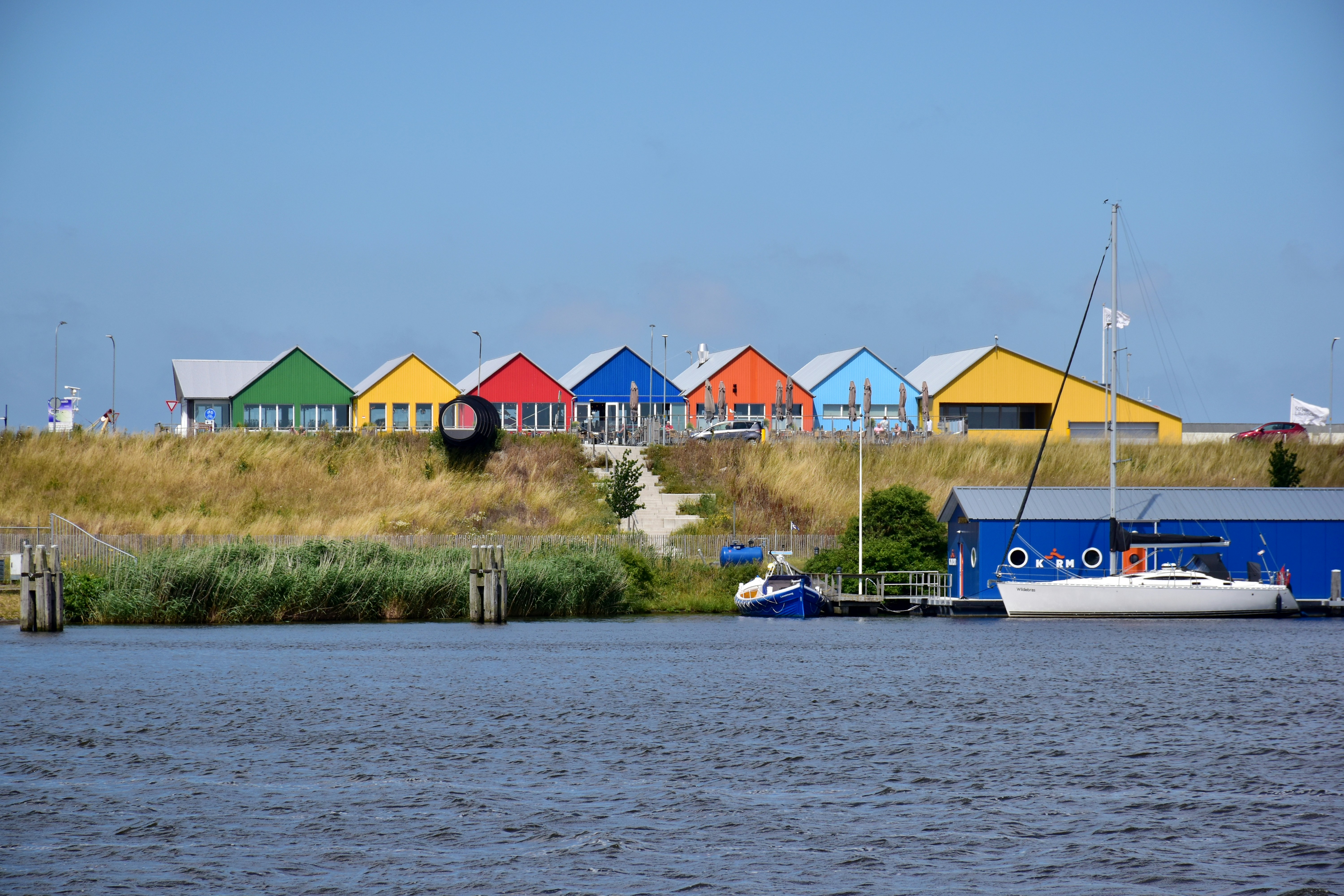Row of colorful houses atop a grassy dike with a yacht in the foreground on a sunny day.