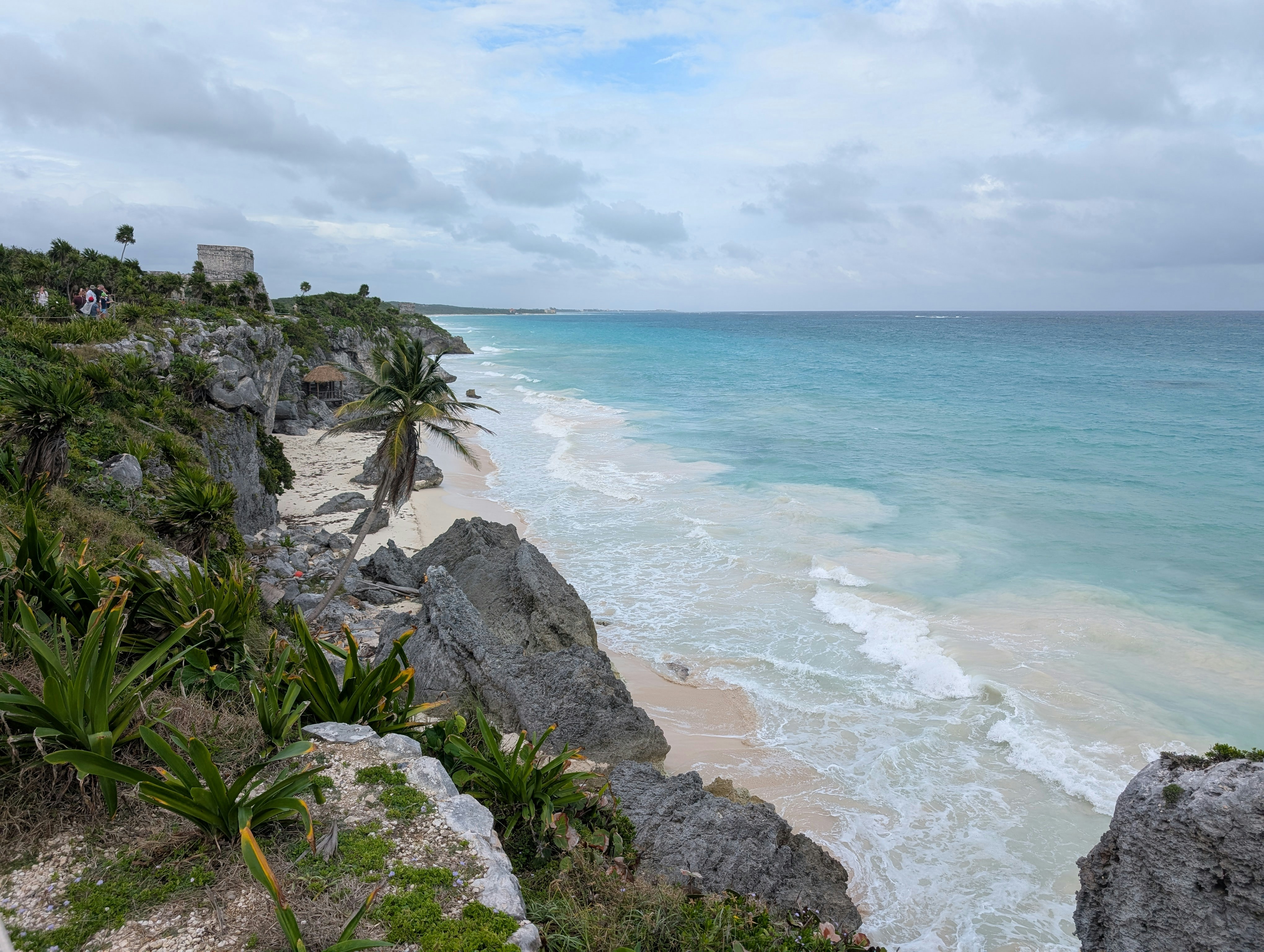 Ancient coastal ruins overlooking vibrant turquoise waters and a sandy beach framed by lush greenery and rugged cliffs.