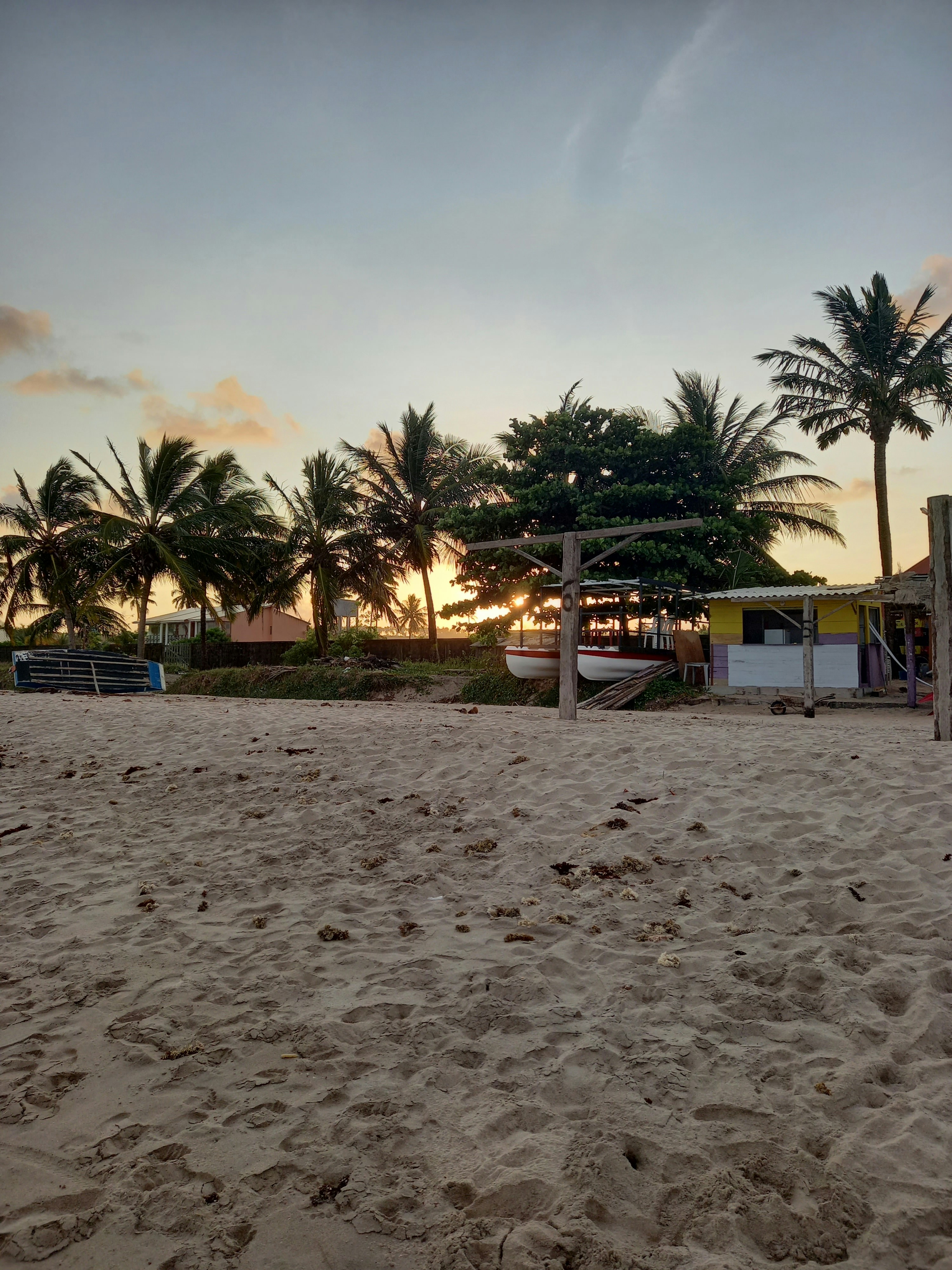 Seaside photograph capturing a palm-fringed beach at sunset with boats on the shore and small huts in the background.