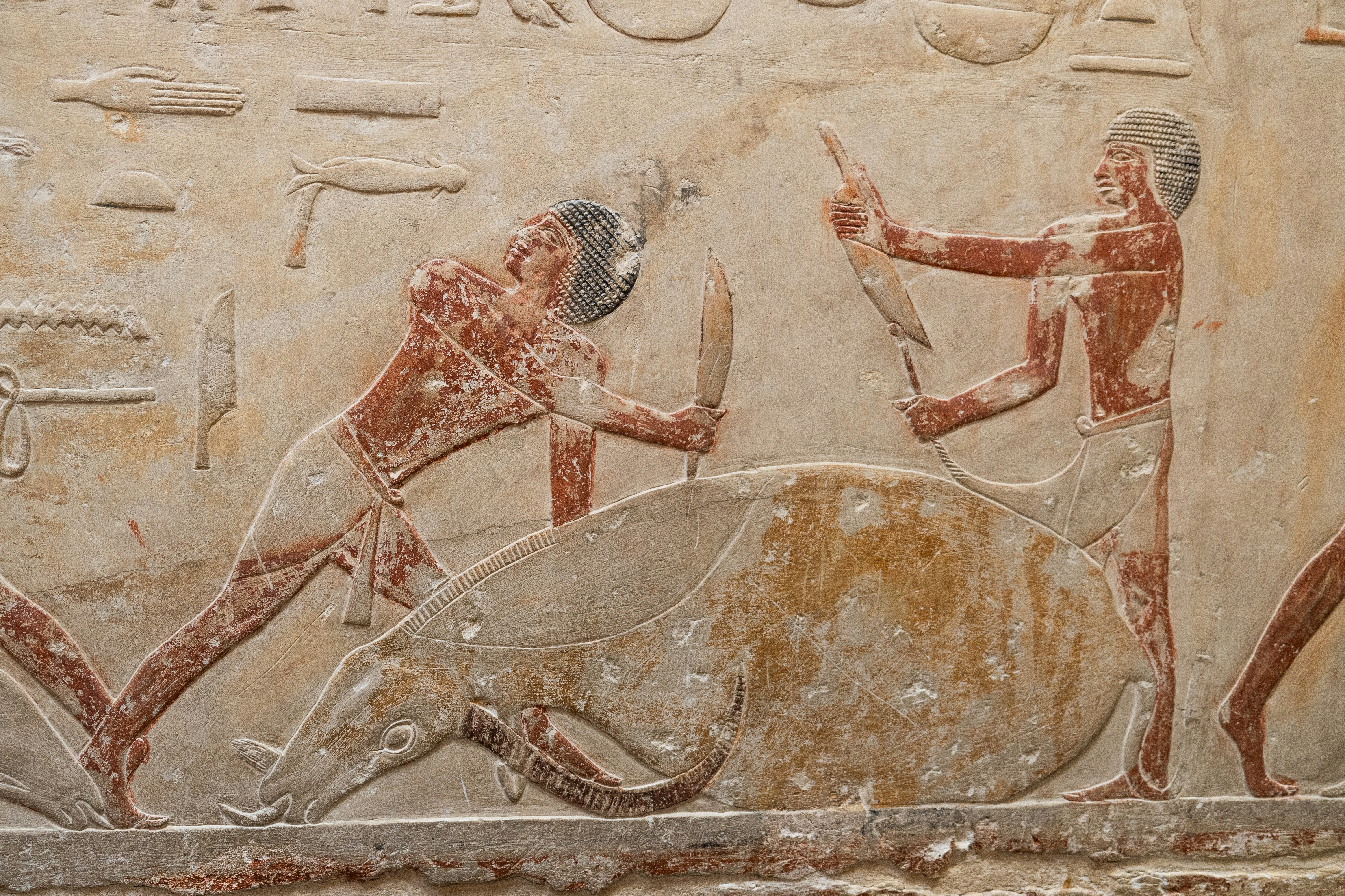 Intricately carved hieroglyphs on a weathered stone wall depict two figures in a ritual with earthy tones and faded pigments.