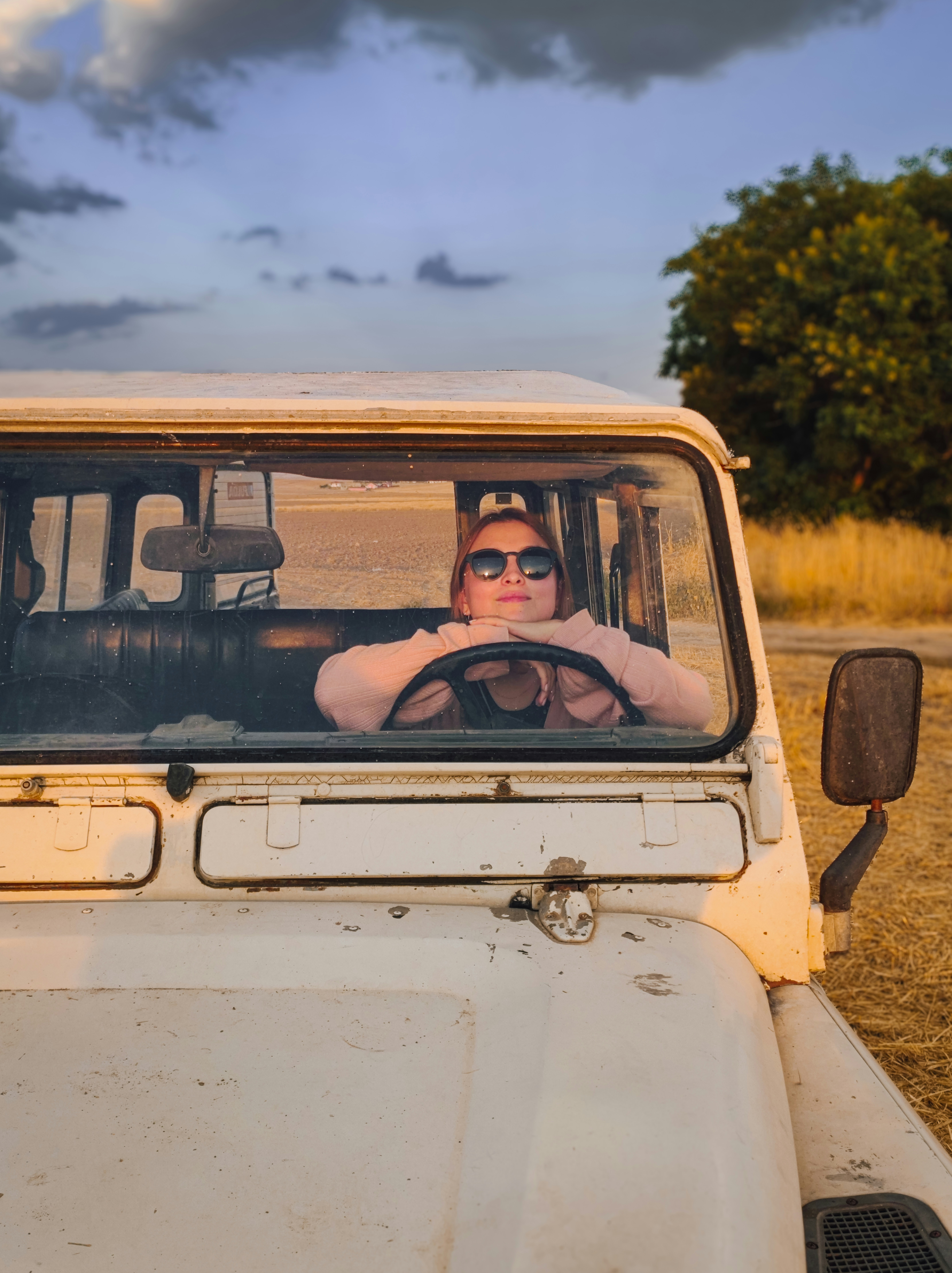 A person wearing sunglasses sits at the wheel of a weathered white jeep, bathed in golden-hour light in a rural field.