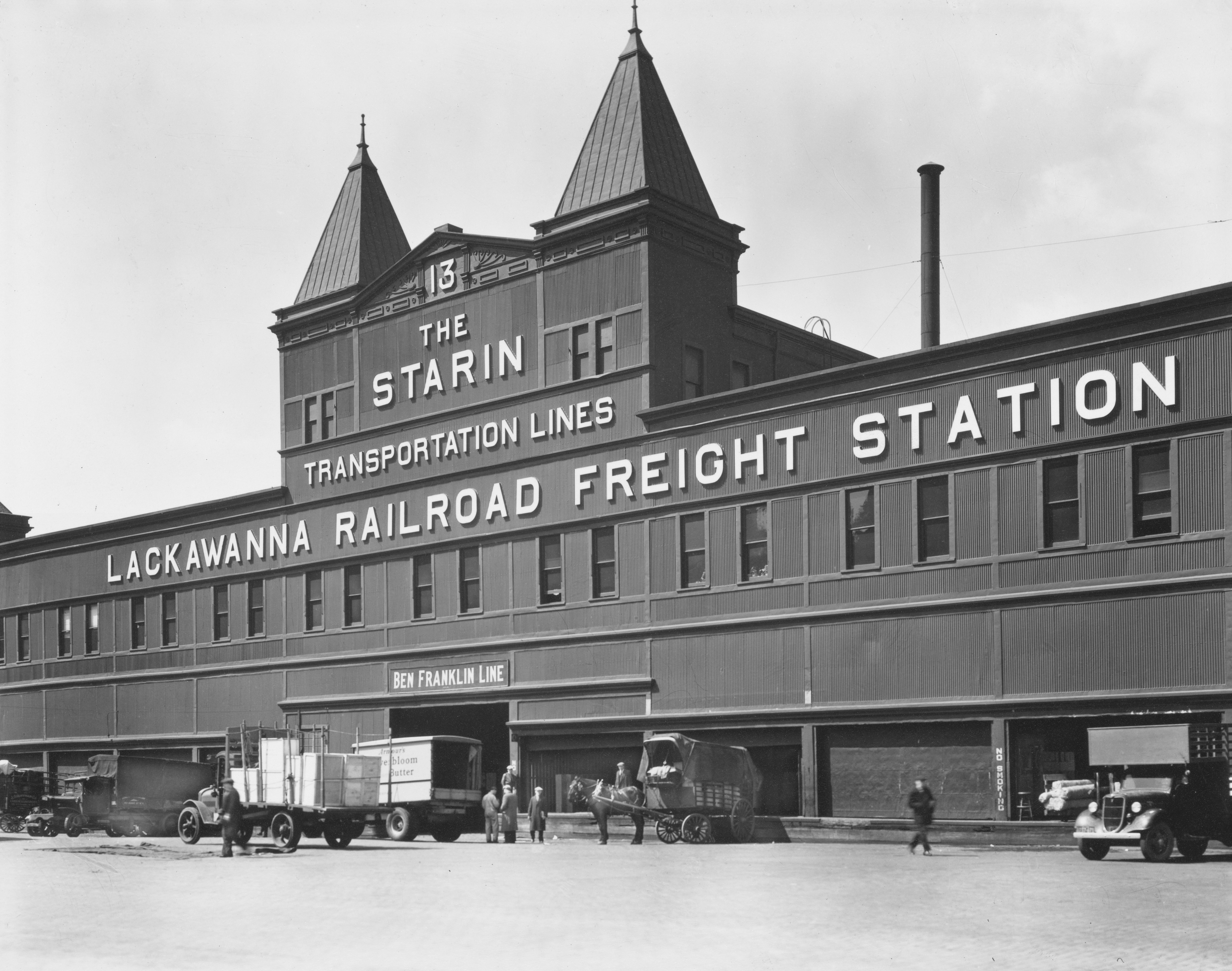 Black and white historic freight station with distinctive pointed towers and bustling activity below.