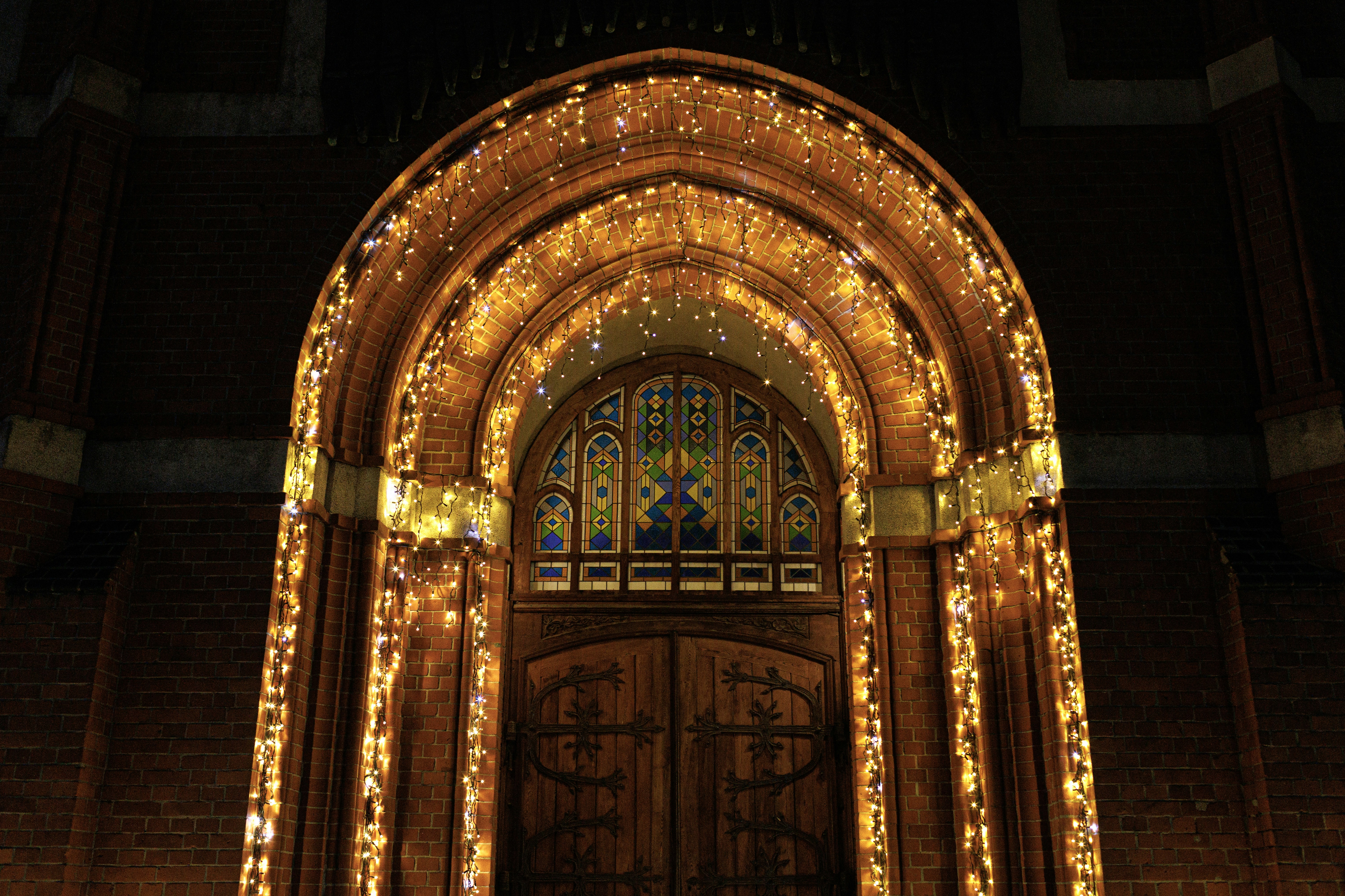 Warm lights drape a brick archway with vibrant stained glass and wooden doors, creating a serene glow.