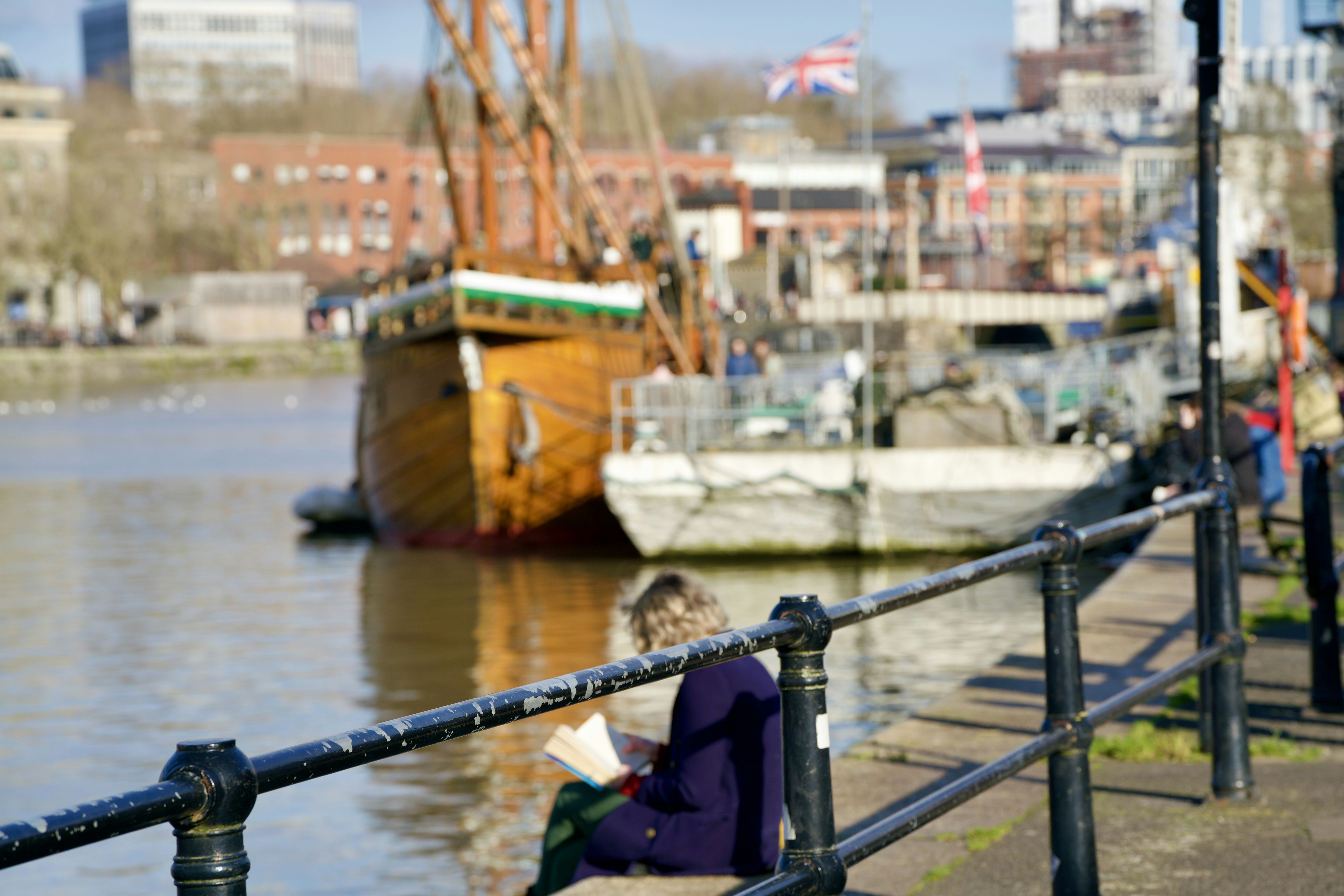 Person reading on a waterfront bench with a historic wooden ship and urban skyline in the background.