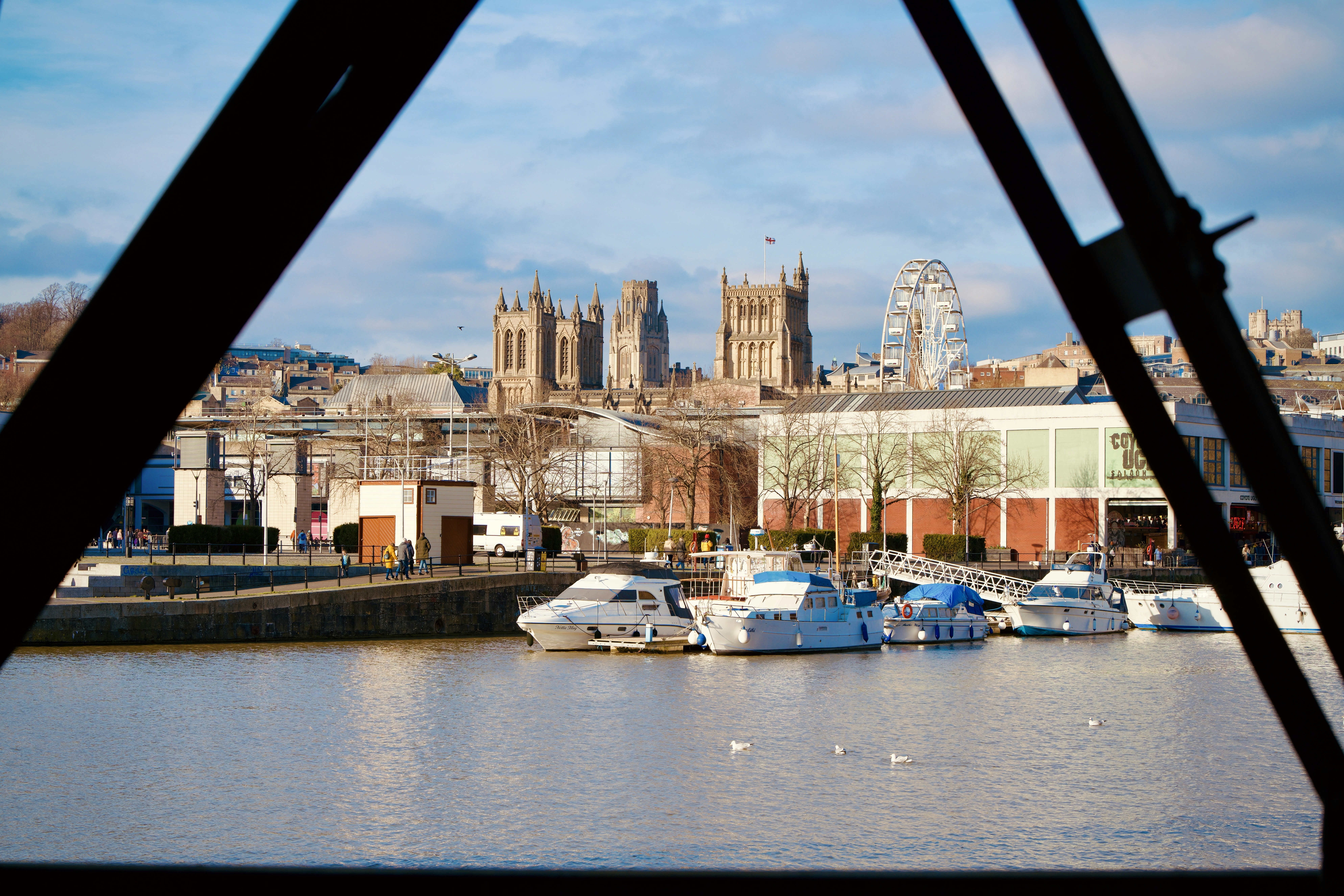Boats docked on a calm river with a historic cathedral and ferris wheel in the background.