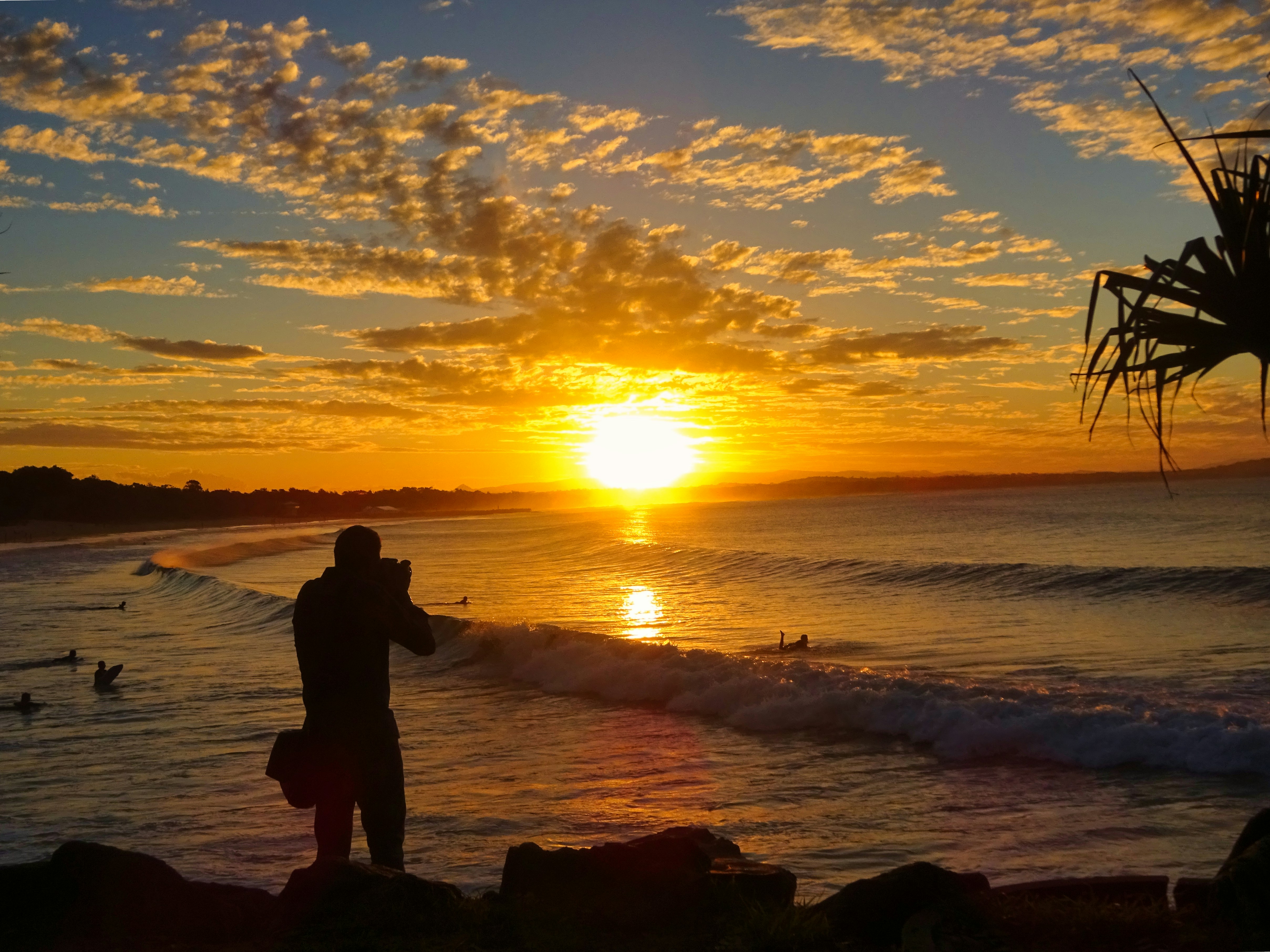 Vibrant sunset over ocean waves with a silhouetted photographer in the foreground and surfers in the glowing water.