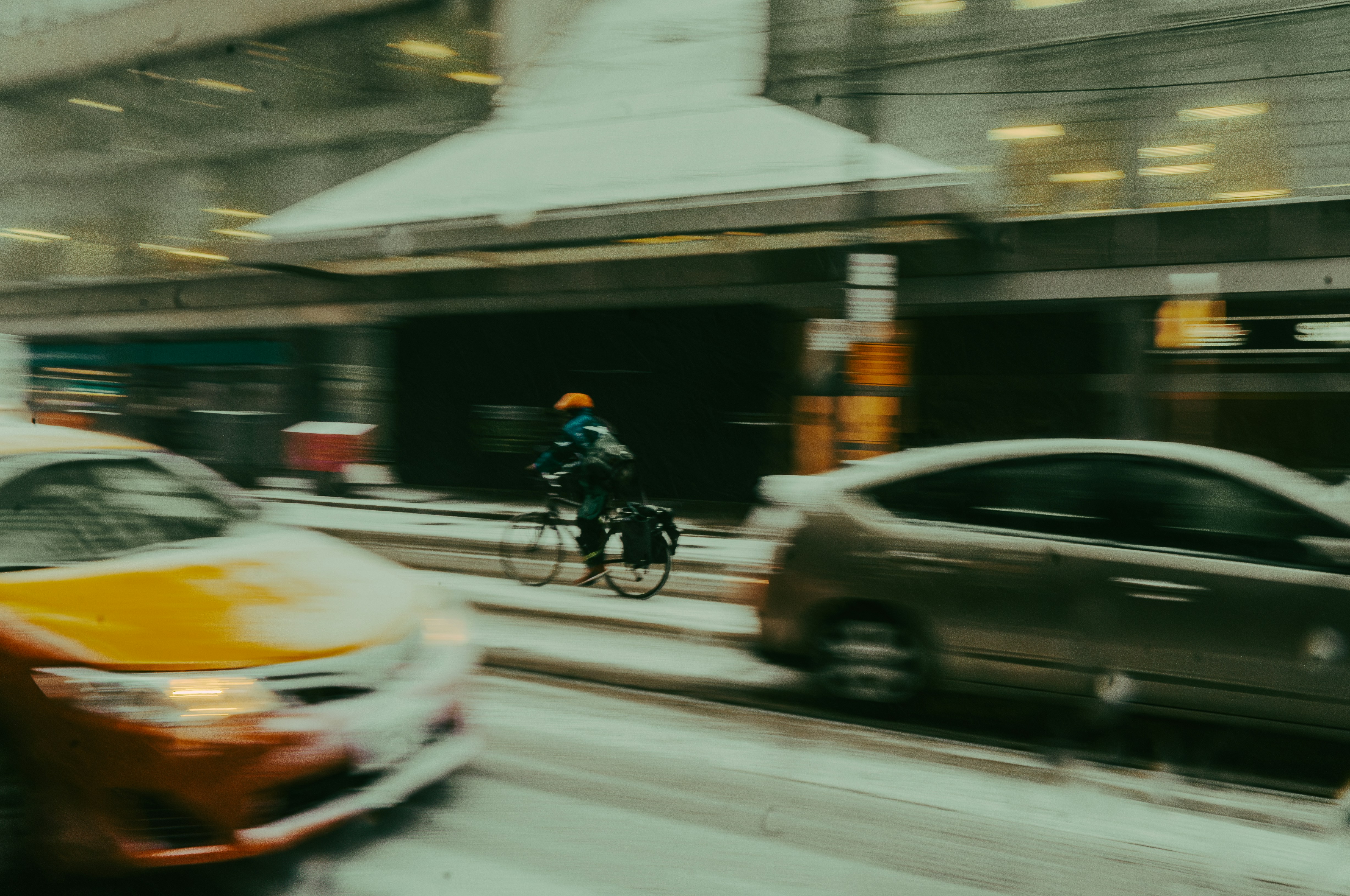 Cyclist with orange helmet rides through snow-covered street amidst blurred traffic, contrasting vibrant colors with muted city tones.