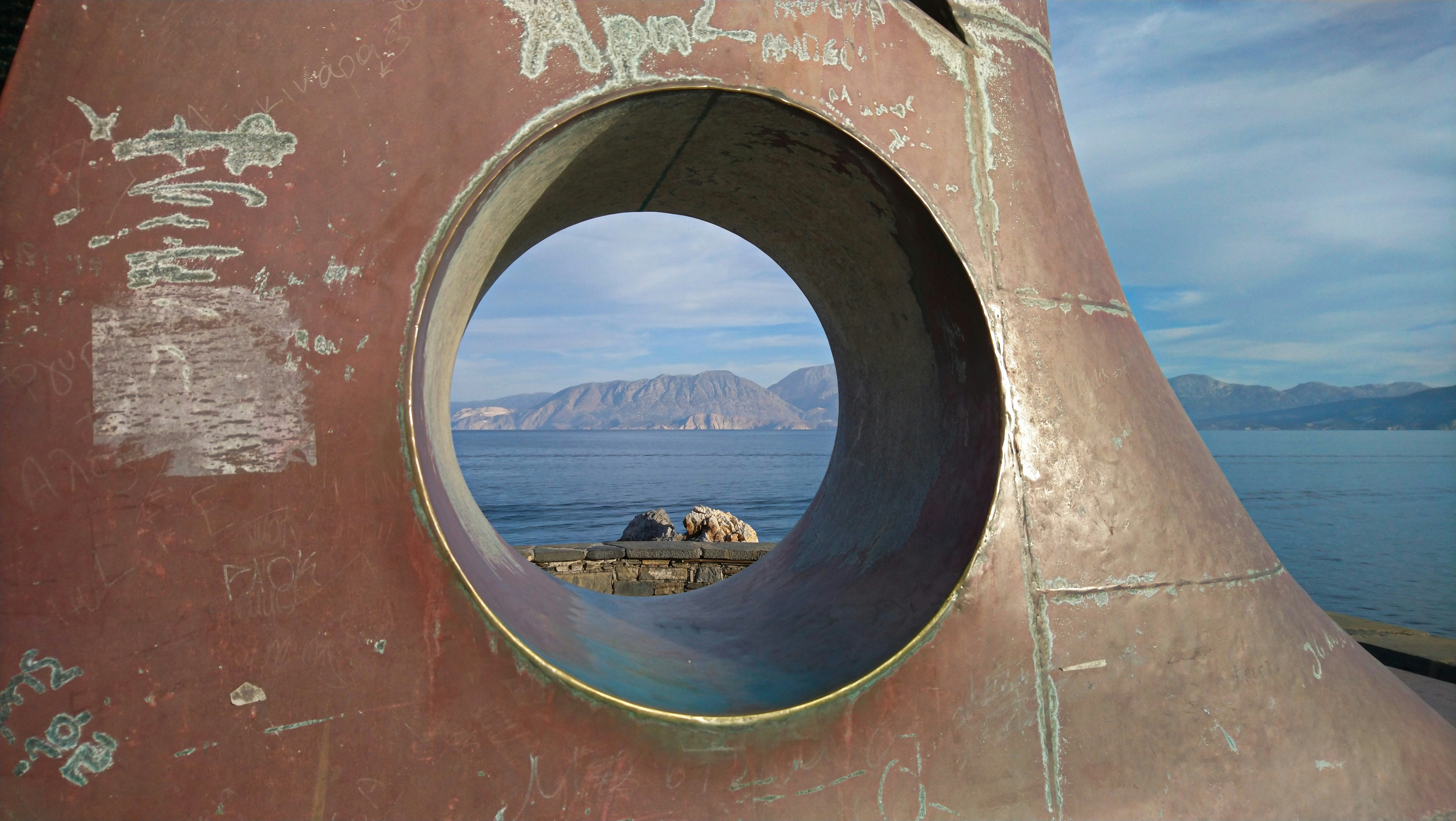 A rusted circular opening frames a calm sea, rocky outcrop, and distant mountains across a clear horizon. The composition contrasts aged metal with the tranquil seascape.