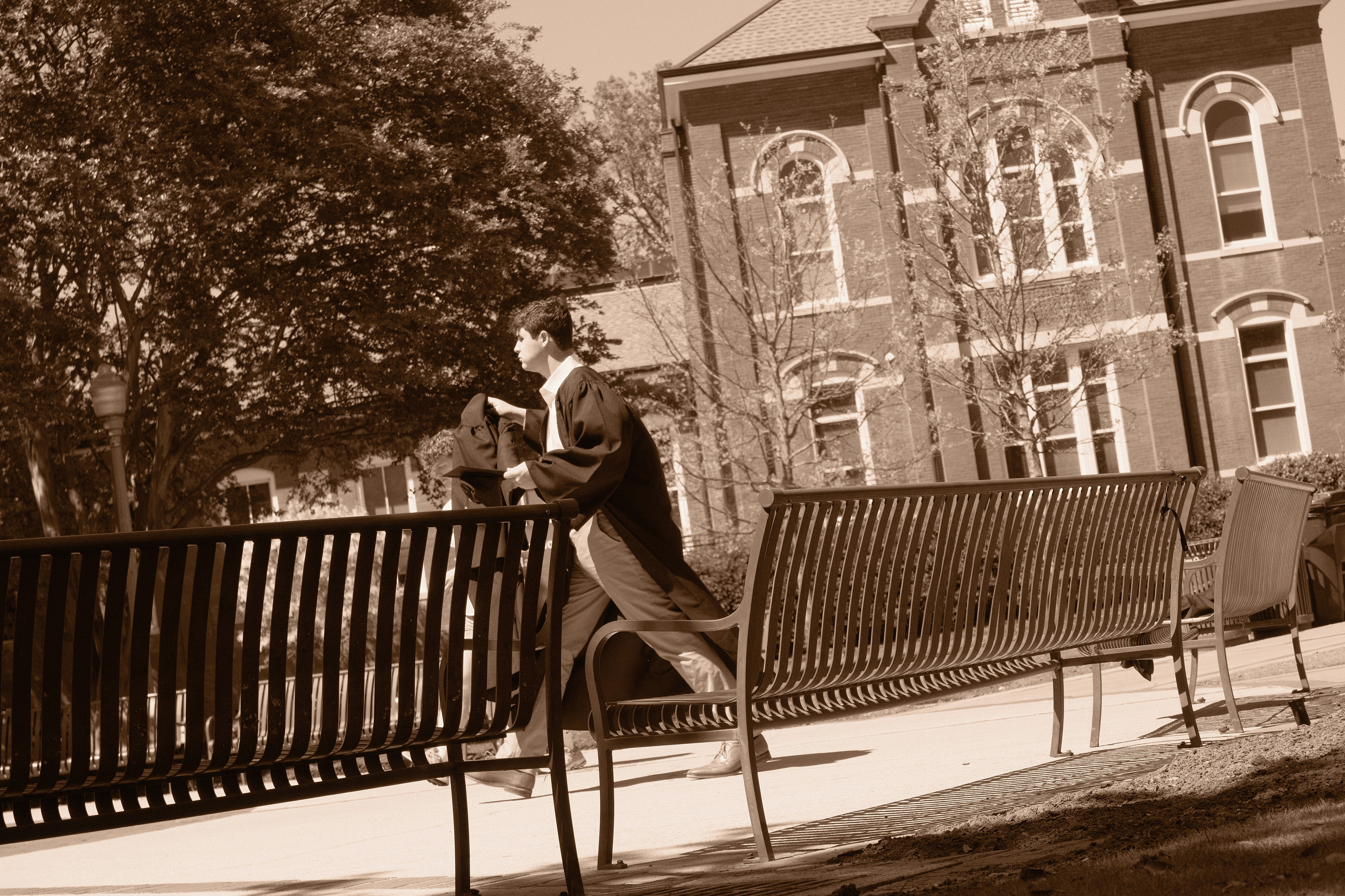 Graduate in flowing gown walks past a classic brick building, framed by benches and trees, in a sepia-toned scene.
