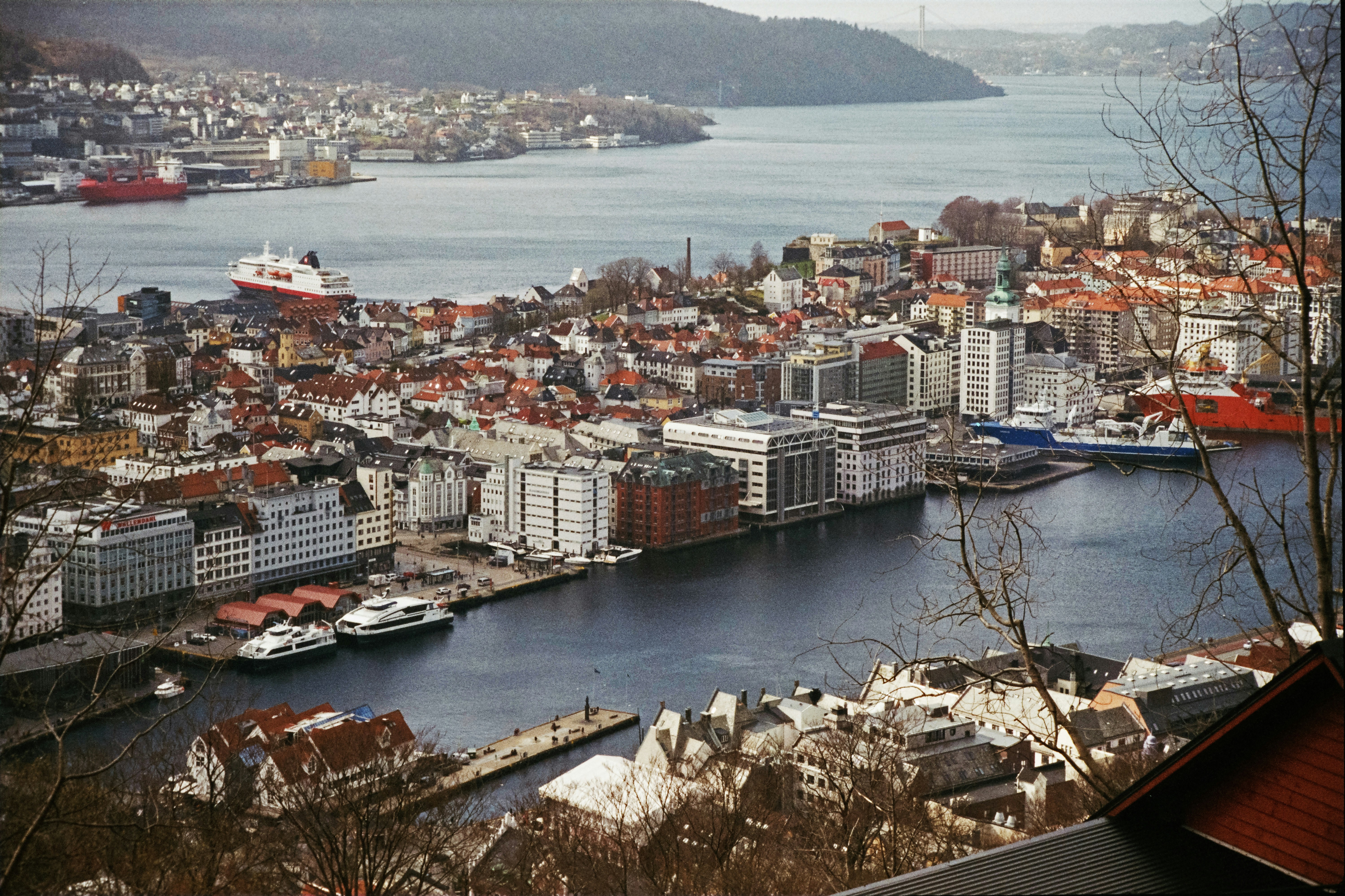 Coastal cityscape with red-roofed buildings and modern architecture by a serene harbor, framed by distant hills.