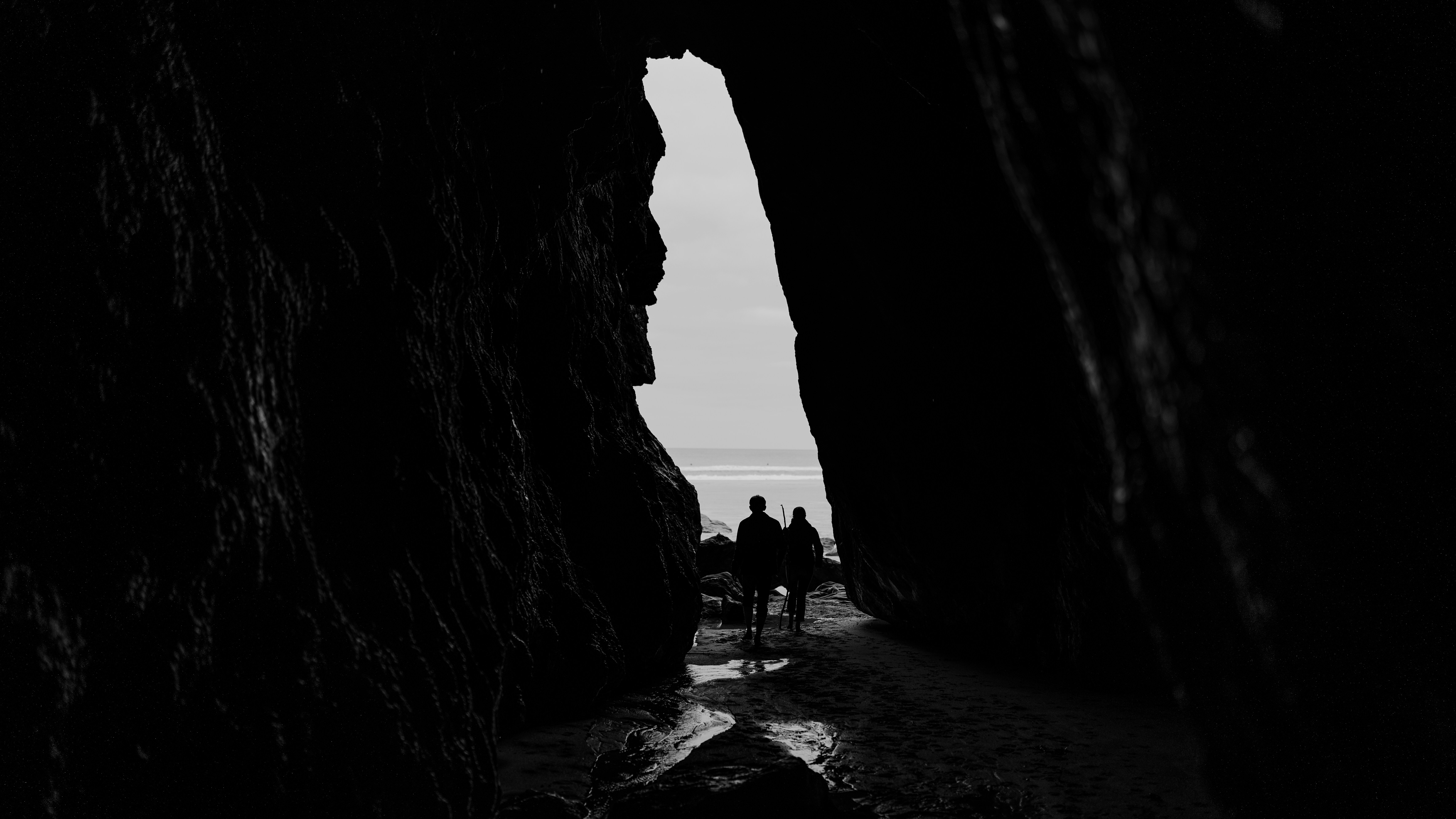 Silhouettes of two people walking through a rugged cave opening towards a distant shoreline. High contrast highlights the texture of the rocks and the reflective path.