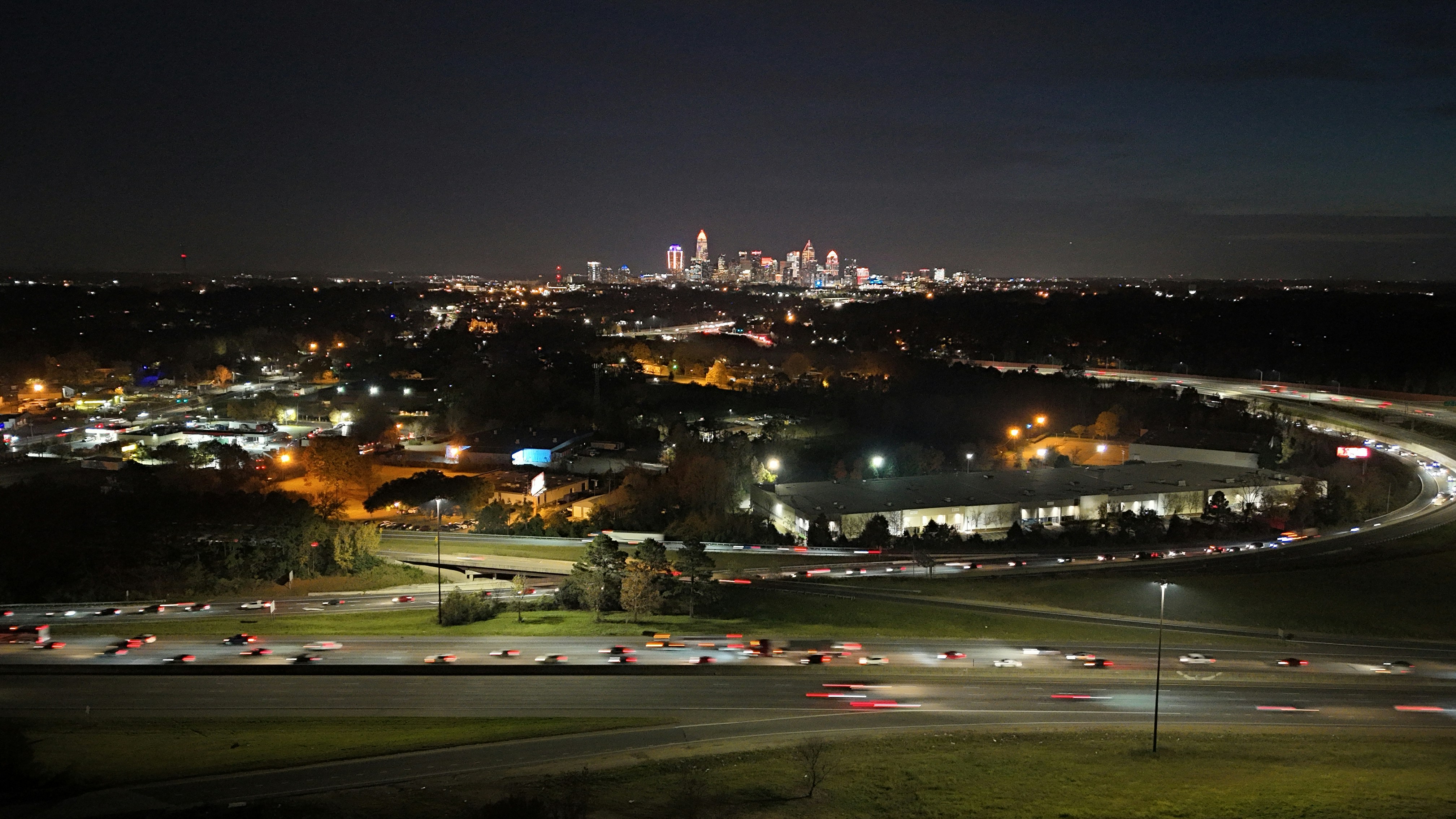 City skyline glowing under a dark sky, with winding roads illuminated by car lights.