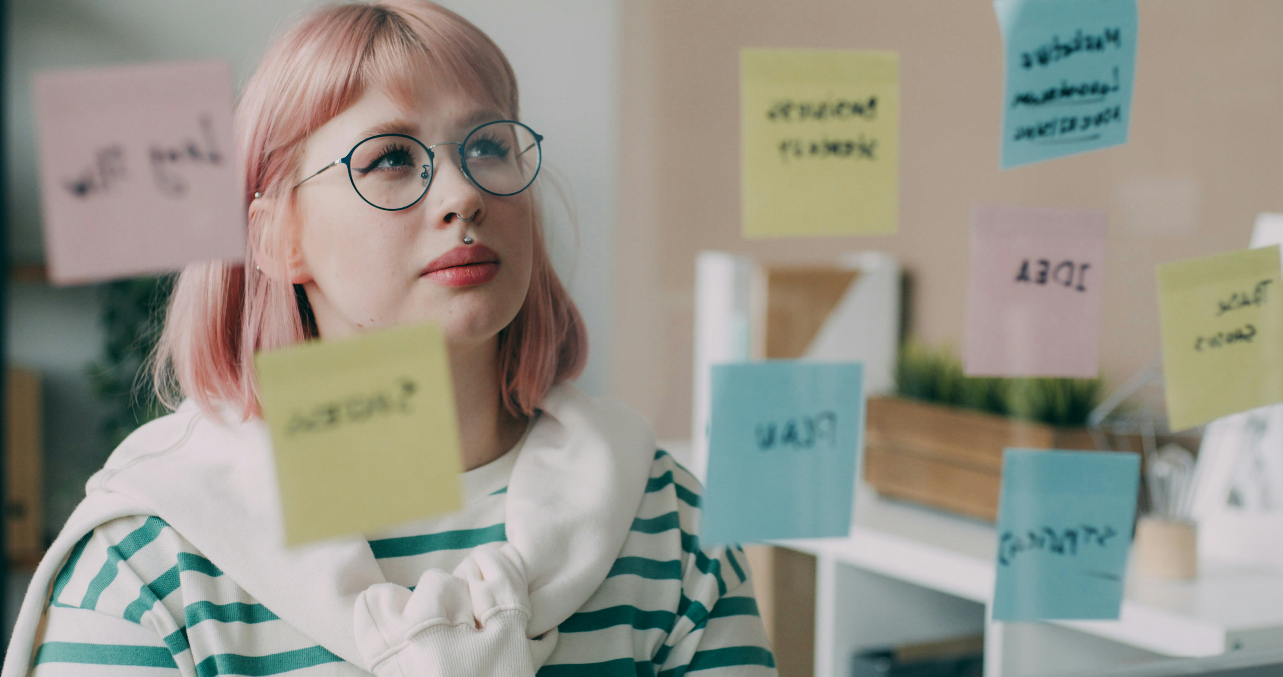 A woman with pink hair and glasses standing in front of post it notes ...