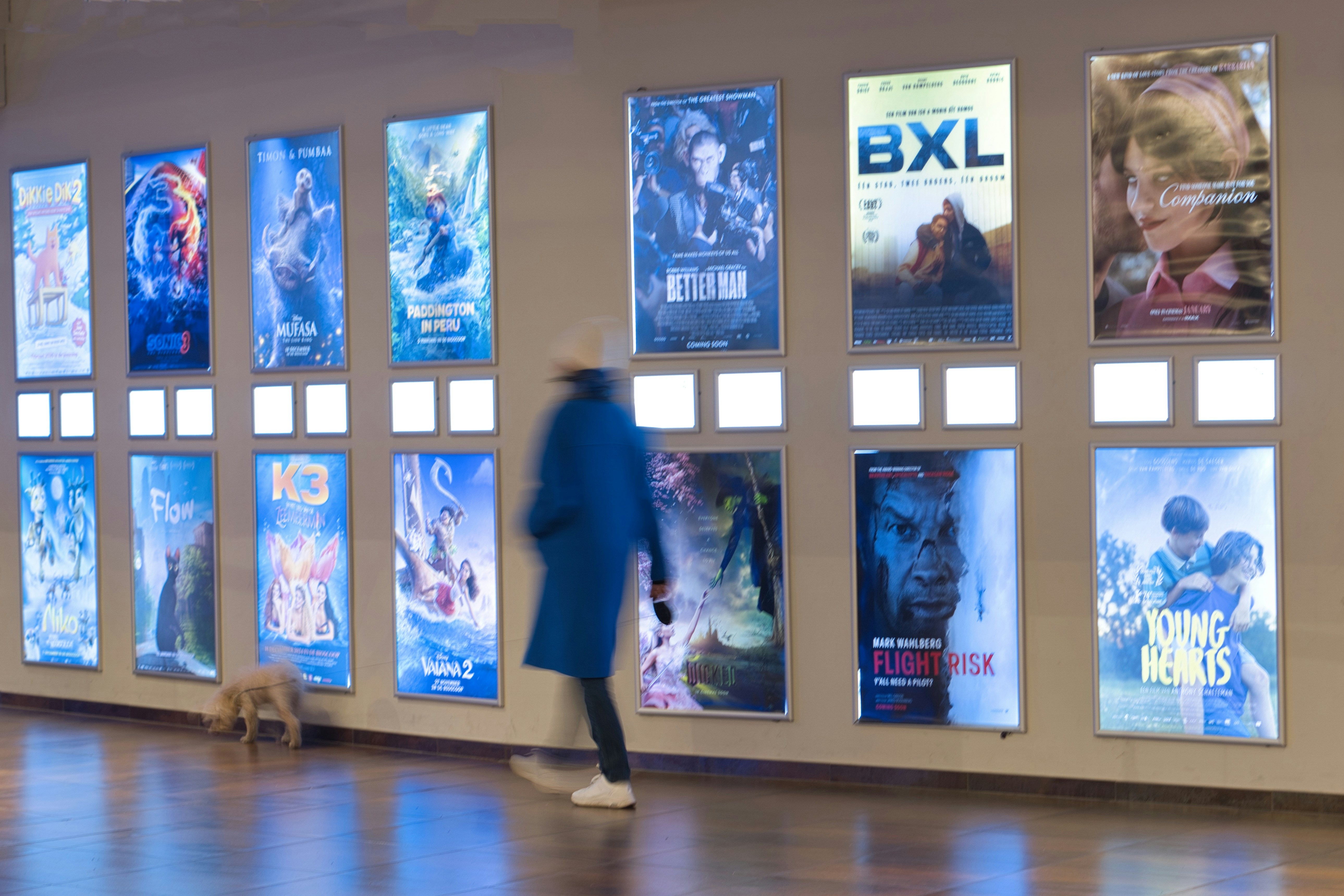 Cinema hallway with vivid movie posters glowing on a neutral wall and a blurred figure in blue walking past.