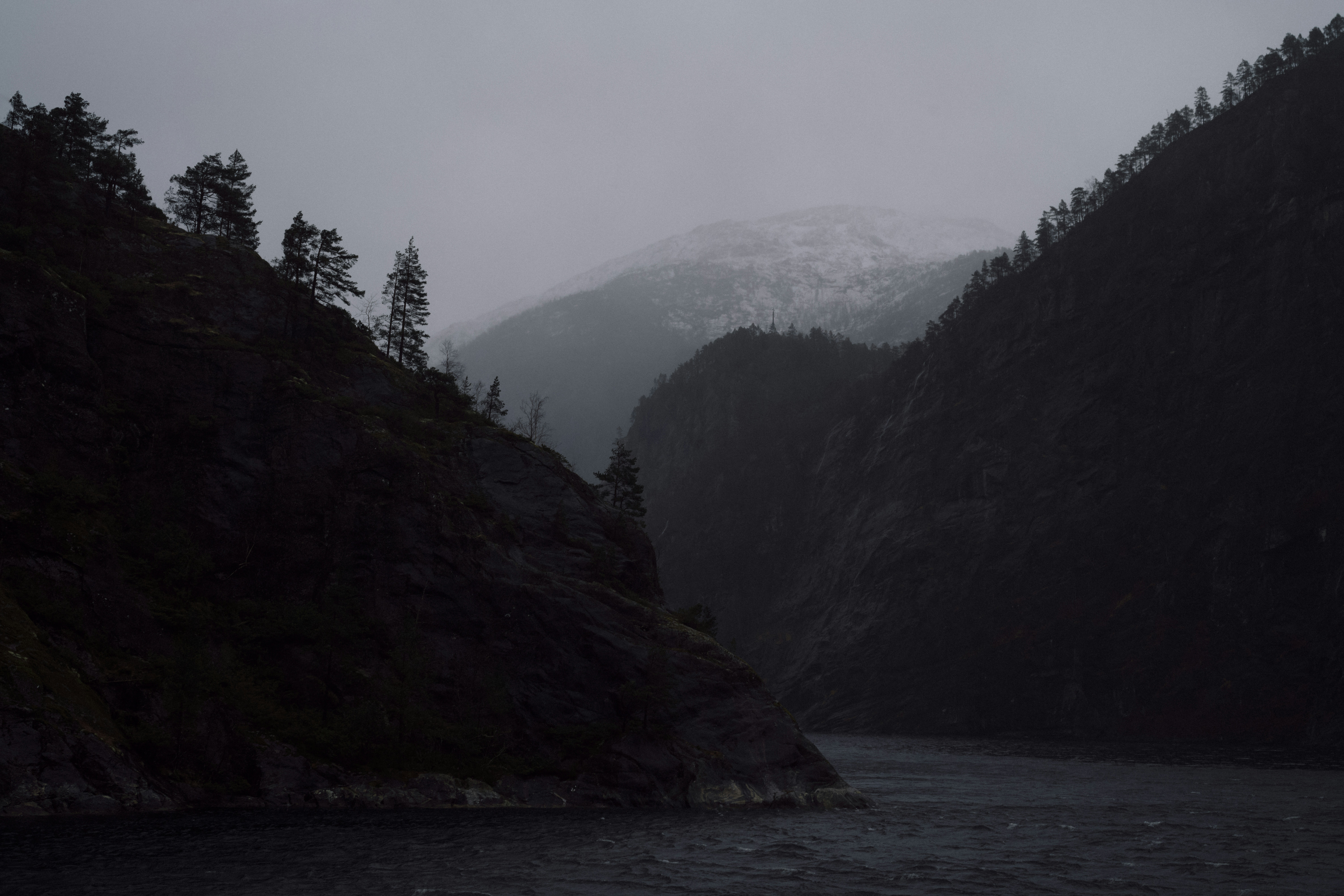 Misty mountain fjord with dark cliffs and sparse trees against a cloudy sky.