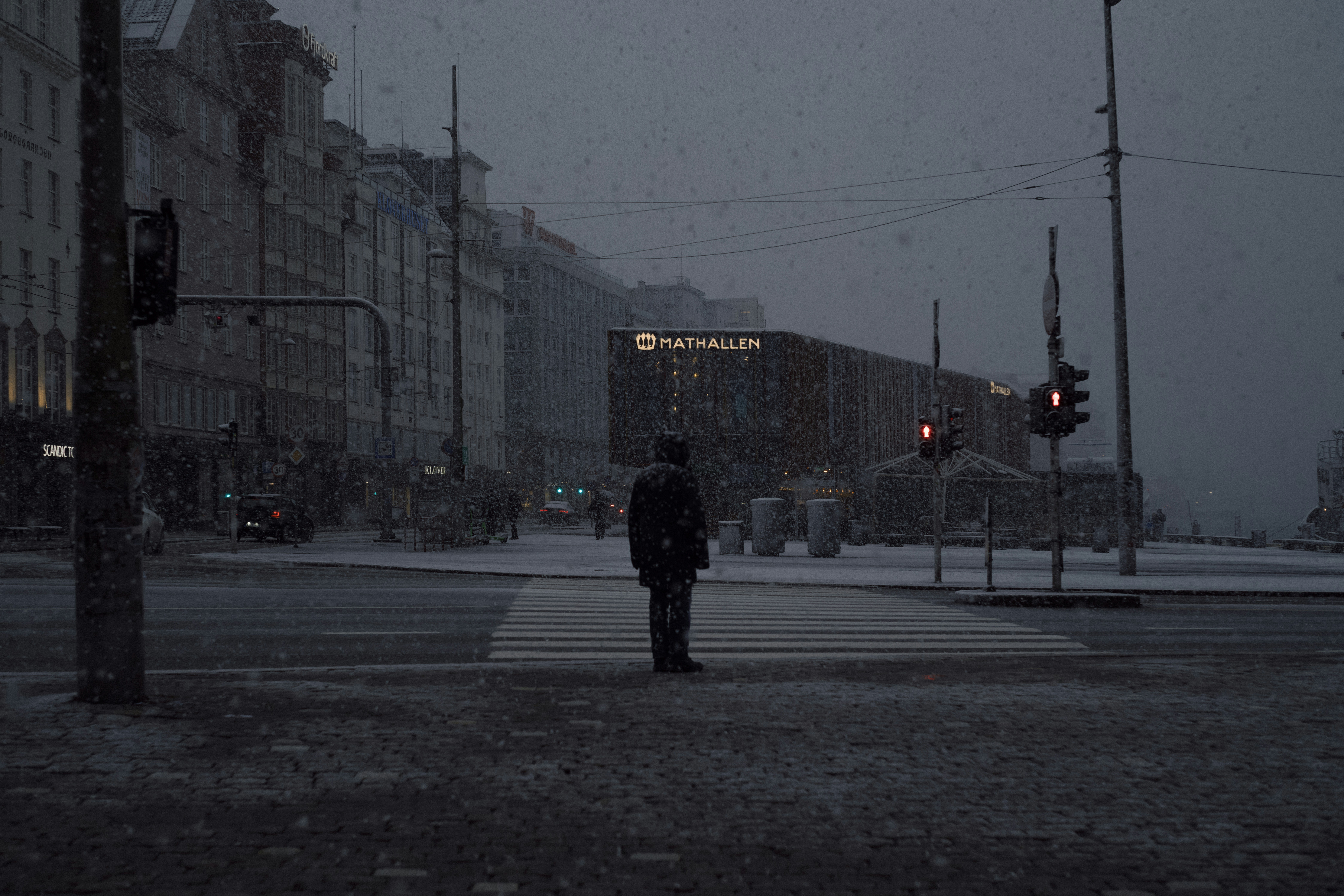 Person standing alone on a snowy city street at dusk with dimly lit buildings in the background.