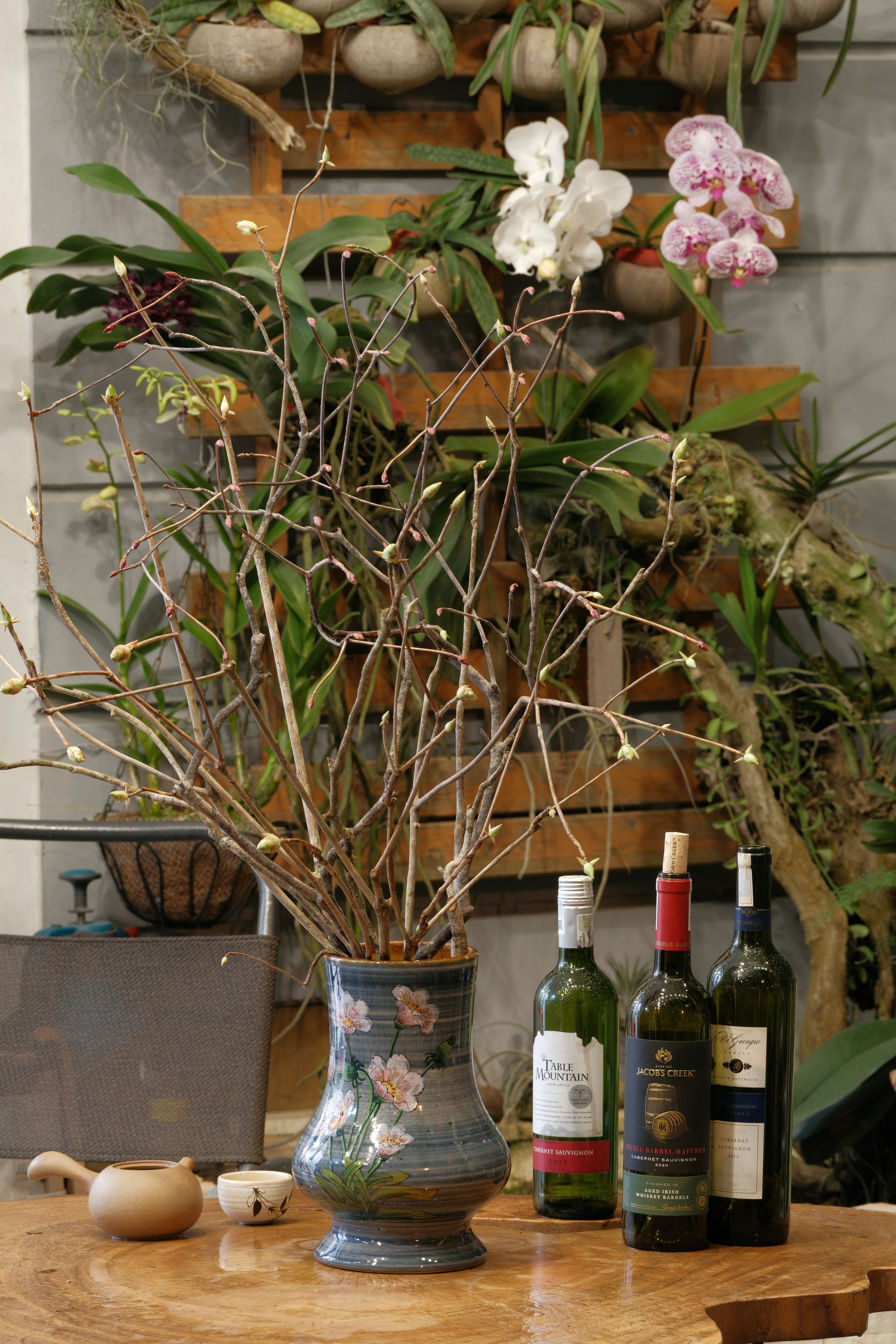 Vase of bare branches crowned by potted orchids and wine bottles on a wooden table