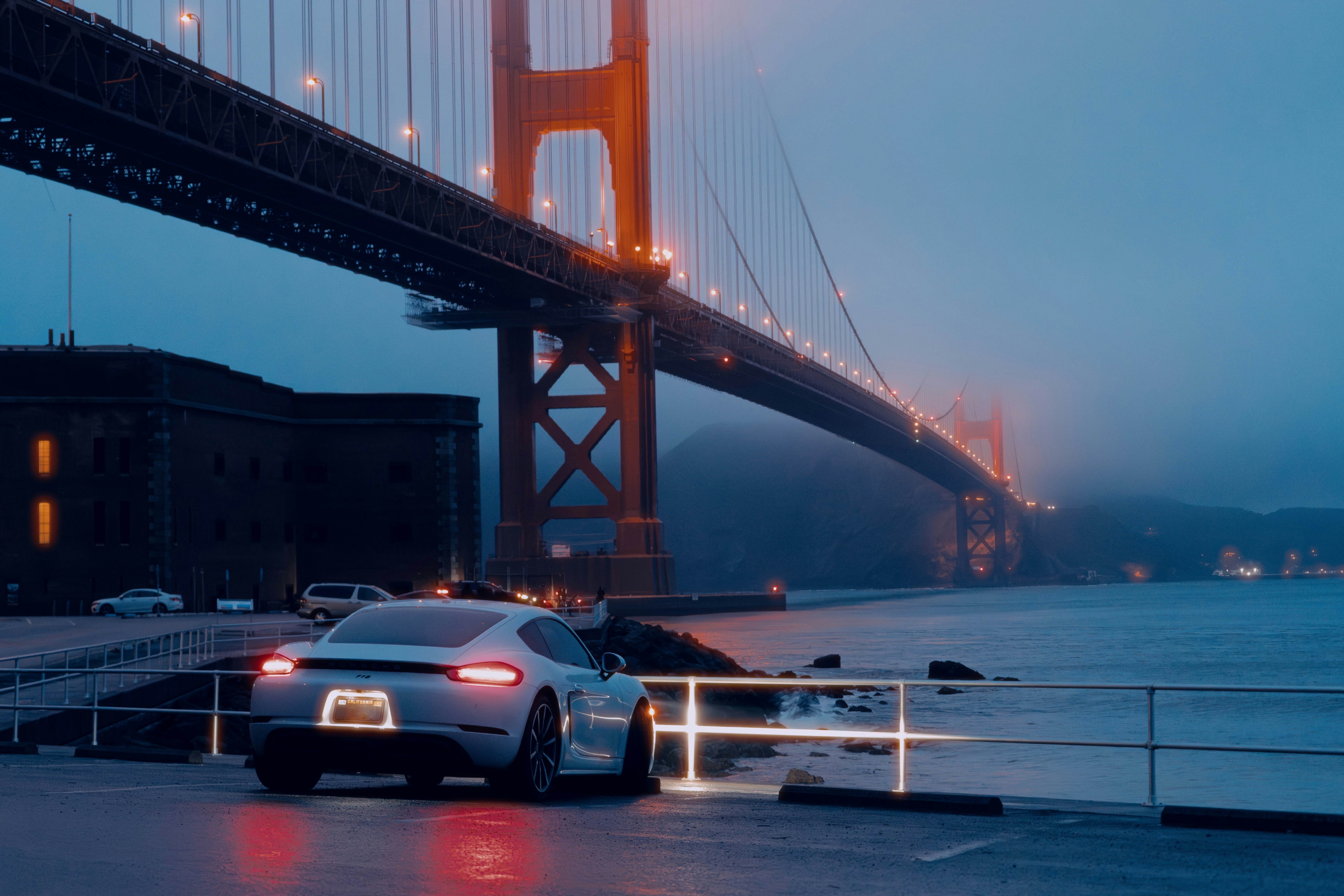White car parked by waterfront under a misty, illuminated bridge at dusk.