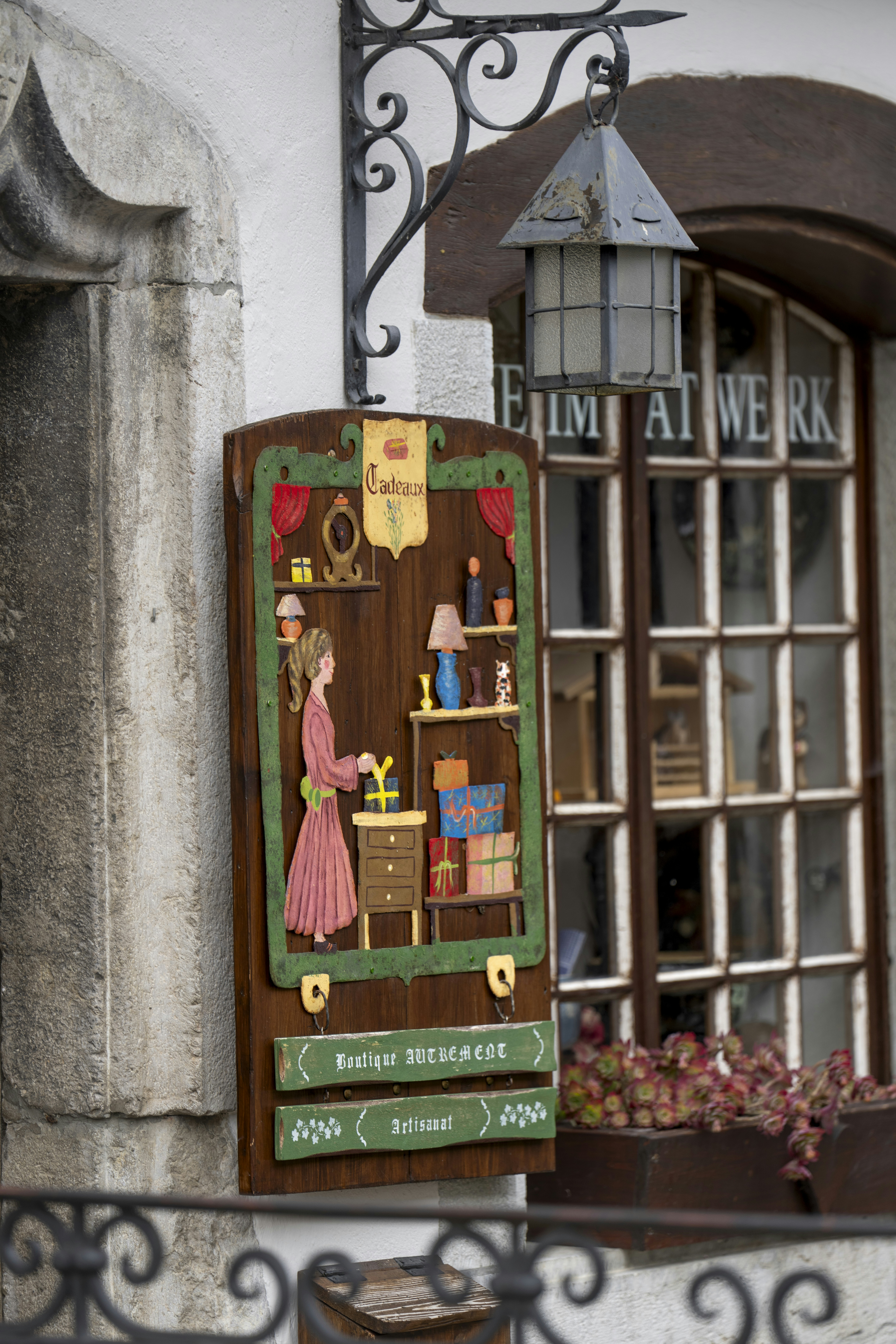 Colorful wooden sign depicting a woman arranging gifts in a quaint shop, showcasing artisanal craftsmanship. A vintage lantern hangs above, adding charm to the scene.