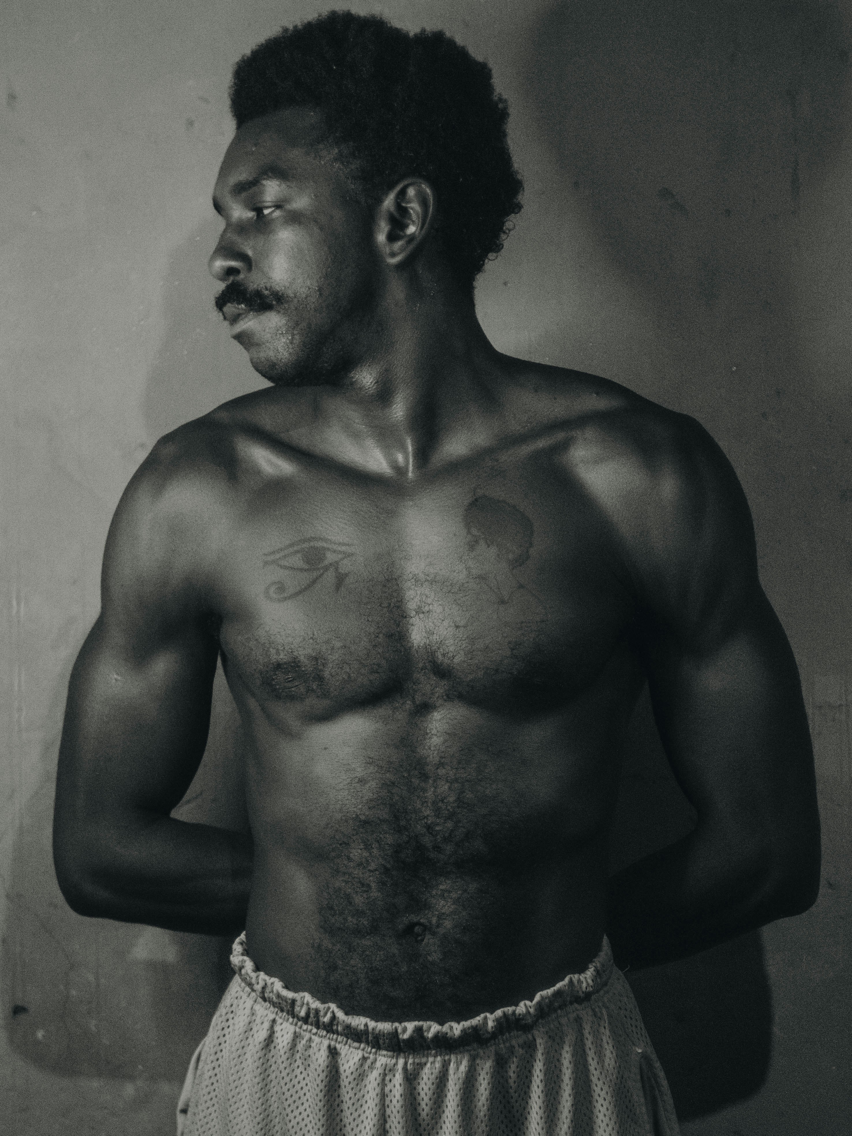 Muscular man with a mustache and tattoos poses against a textured wall, exuding confidence and calmness.