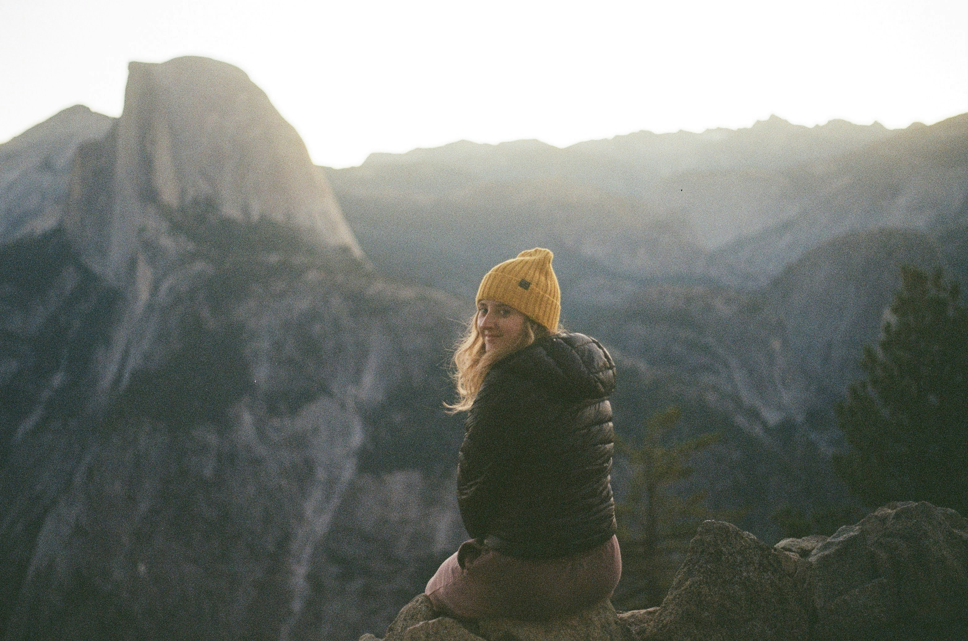 Woman in yellow beanie sits on rocky ledge overlooking Half Dome at sunset.