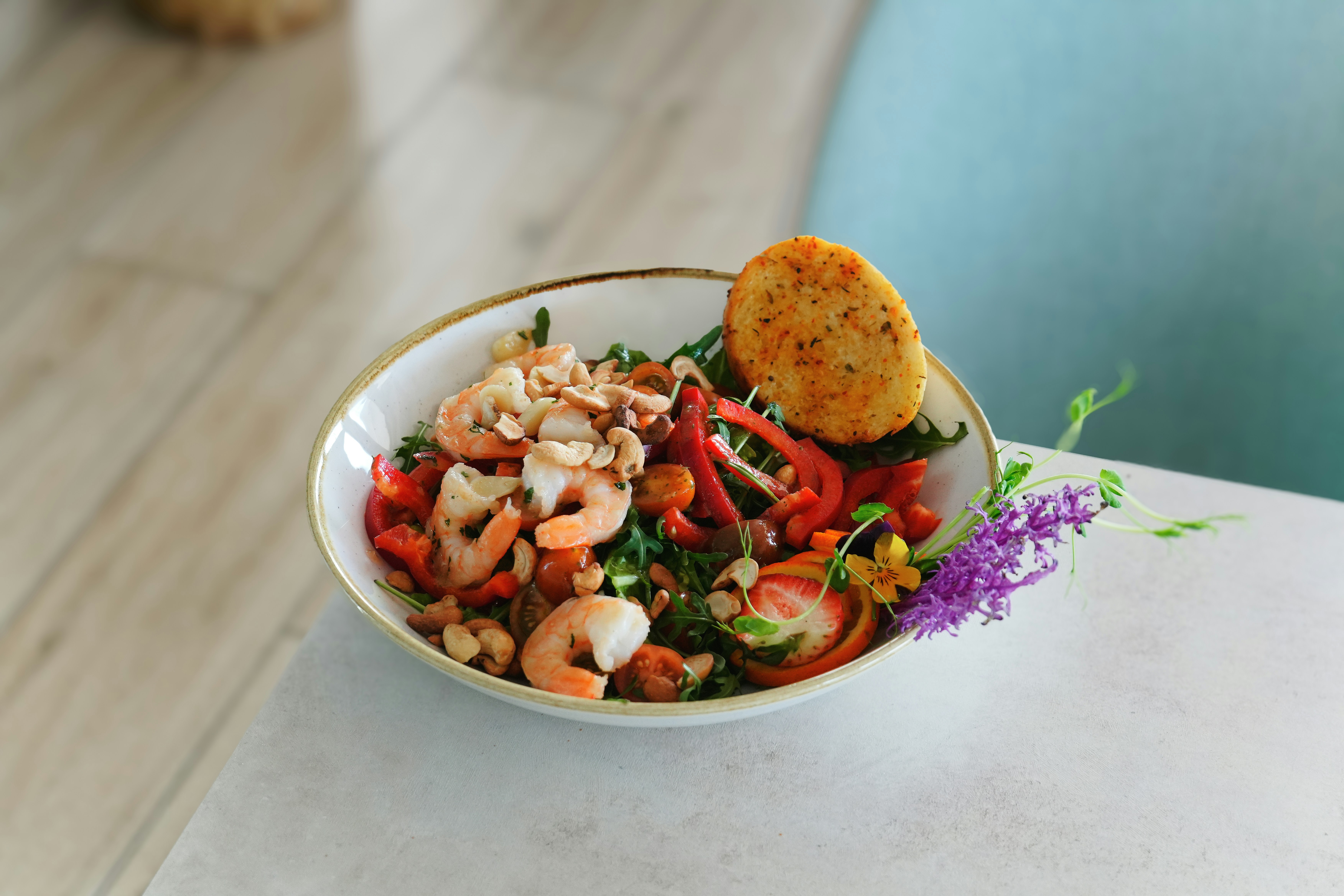 Seafood salad with shrimp, nuts, red bell peppers, and greens, garnished with crostini and flowers on a light background.