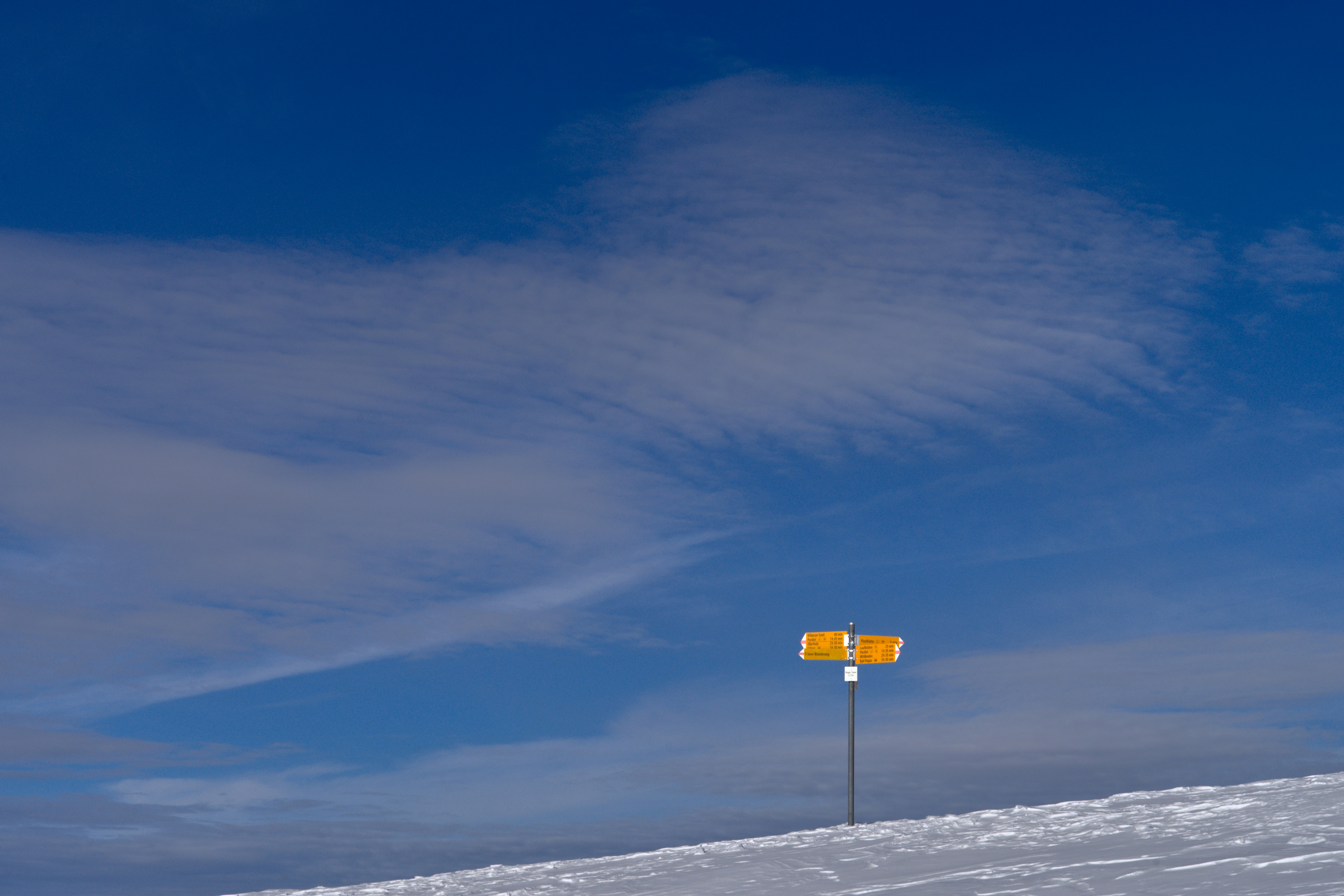 Yellow signpost stands on snowy slope beneath a deep blue sky with wispy clouds.