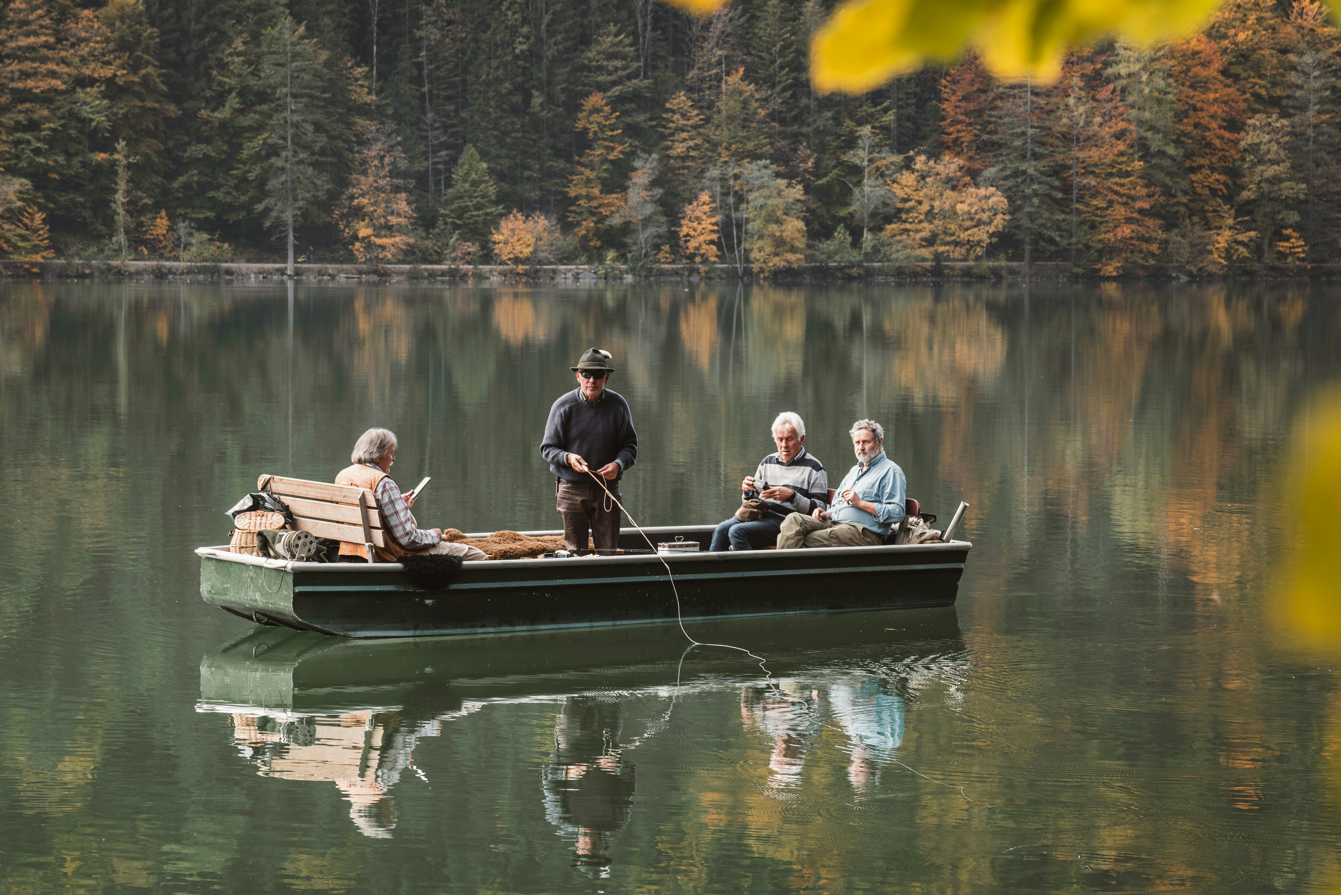 Four people fishing on a small boat in a calm lake, surrounded by vibrant autumn trees and their reflections.