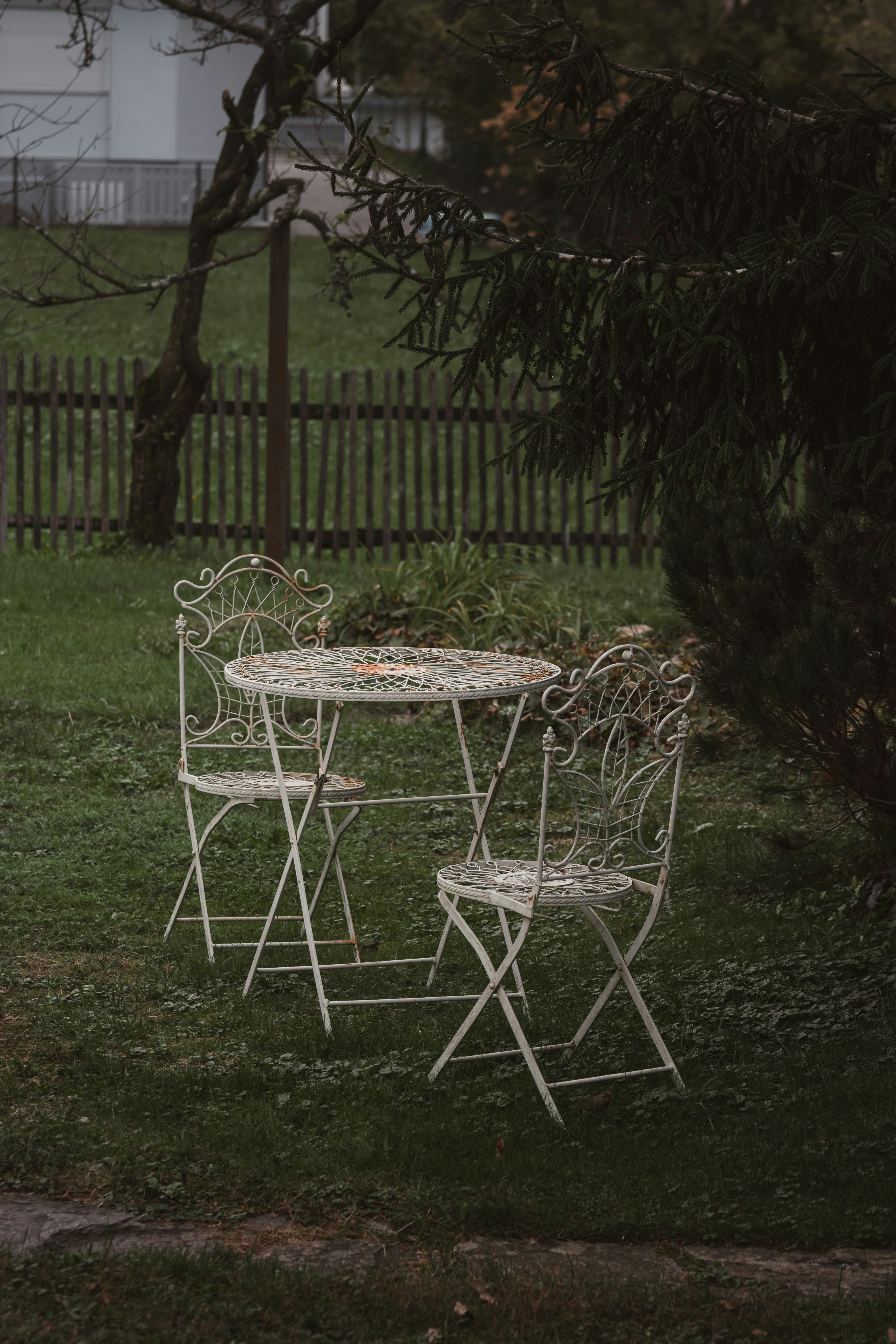 White wrought-iron table with two matching chairs sits on a grassy garden, with a wooden fence and trees in the background.