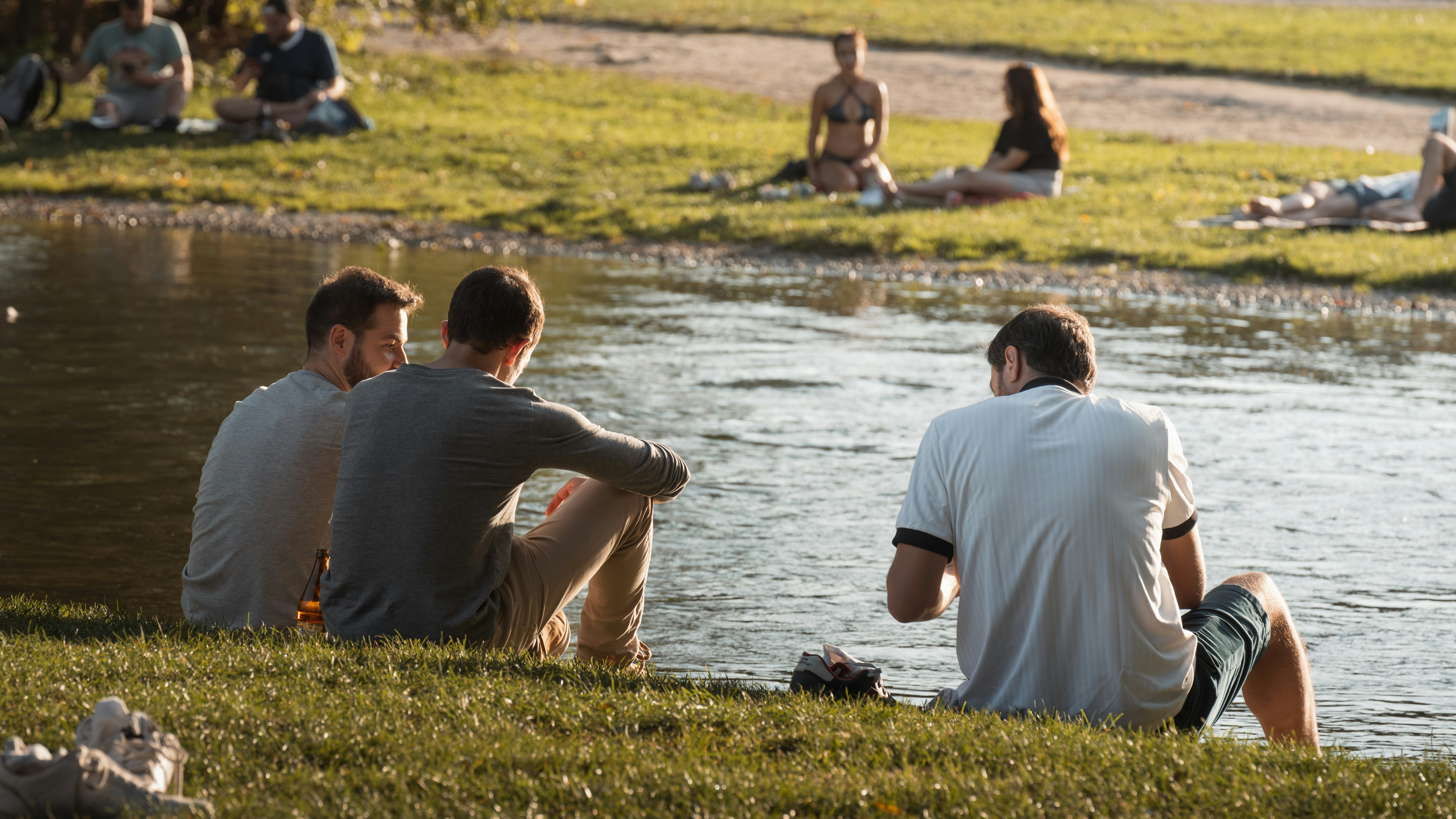 People relaxing by a riverside, bathed in warm sunlight with vibrant green grass and gentle water ripples.