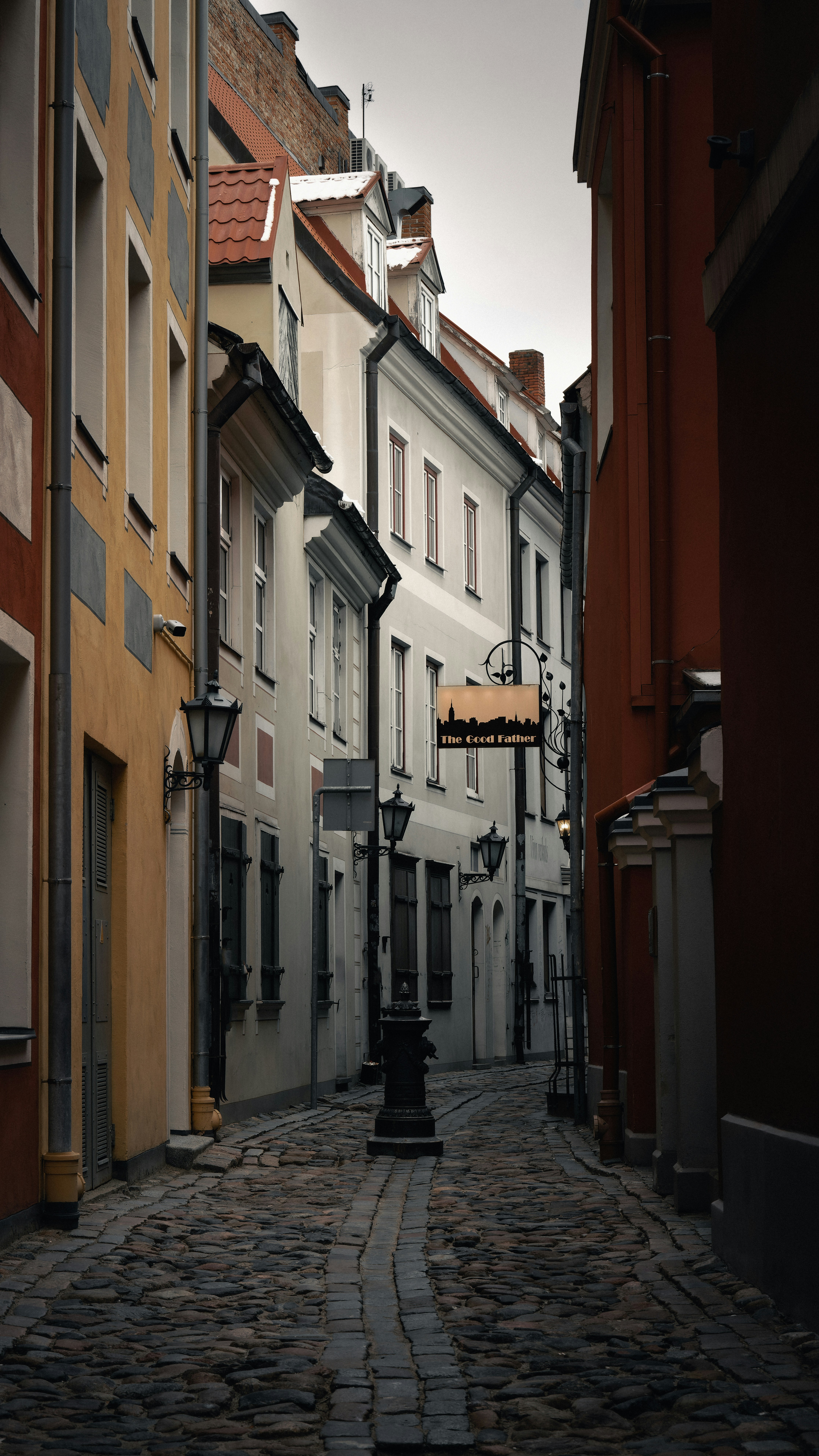 Charming cobblestone alley flanked by colorful buildings, featuring vintage street lamps and a sign for 'The Good Father.'