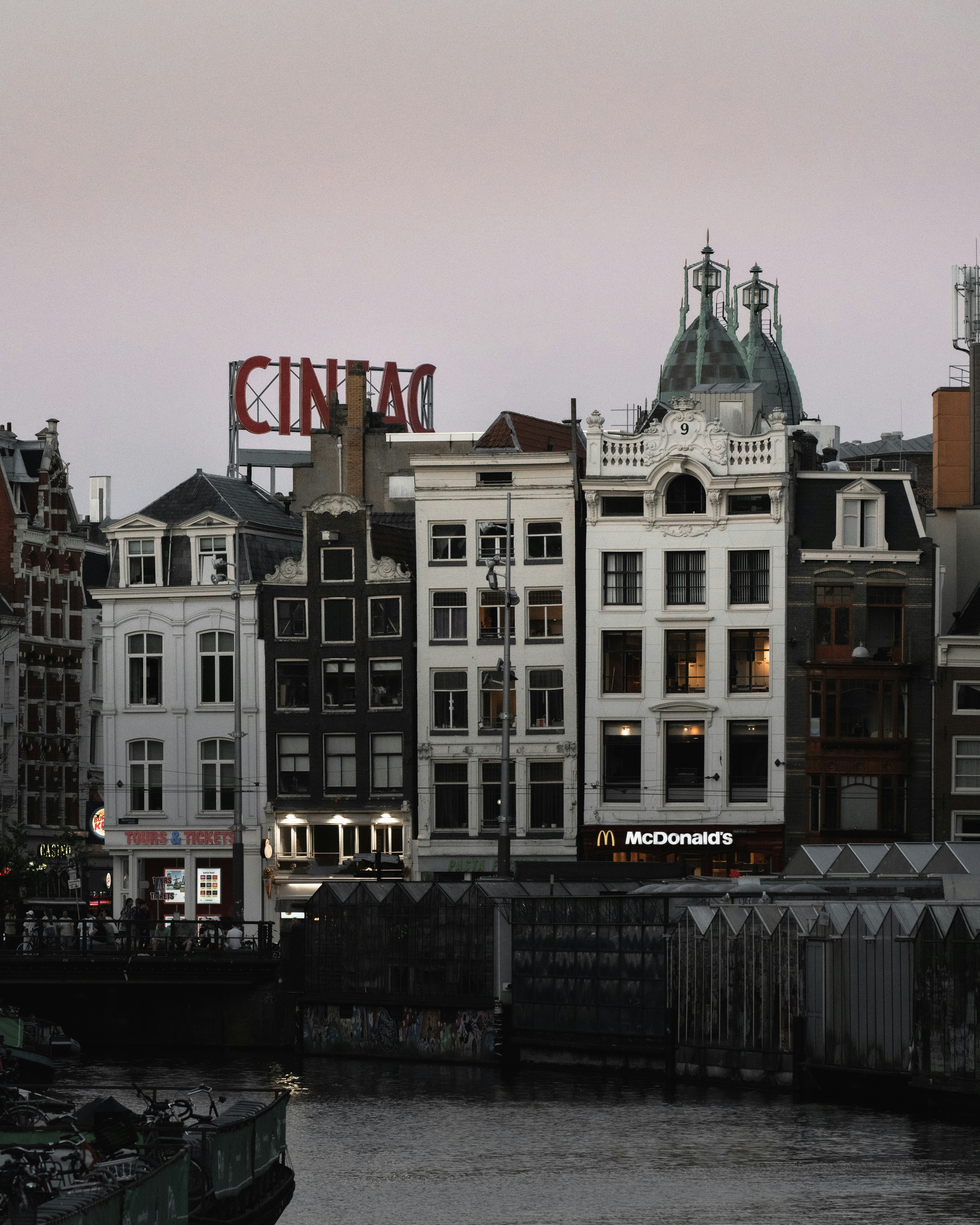 Historic buildings lining a canal in Amsterdam, showcasing a mix of traditional and modern elements. The iconic CINTAC sign adds a nostalgic touch.