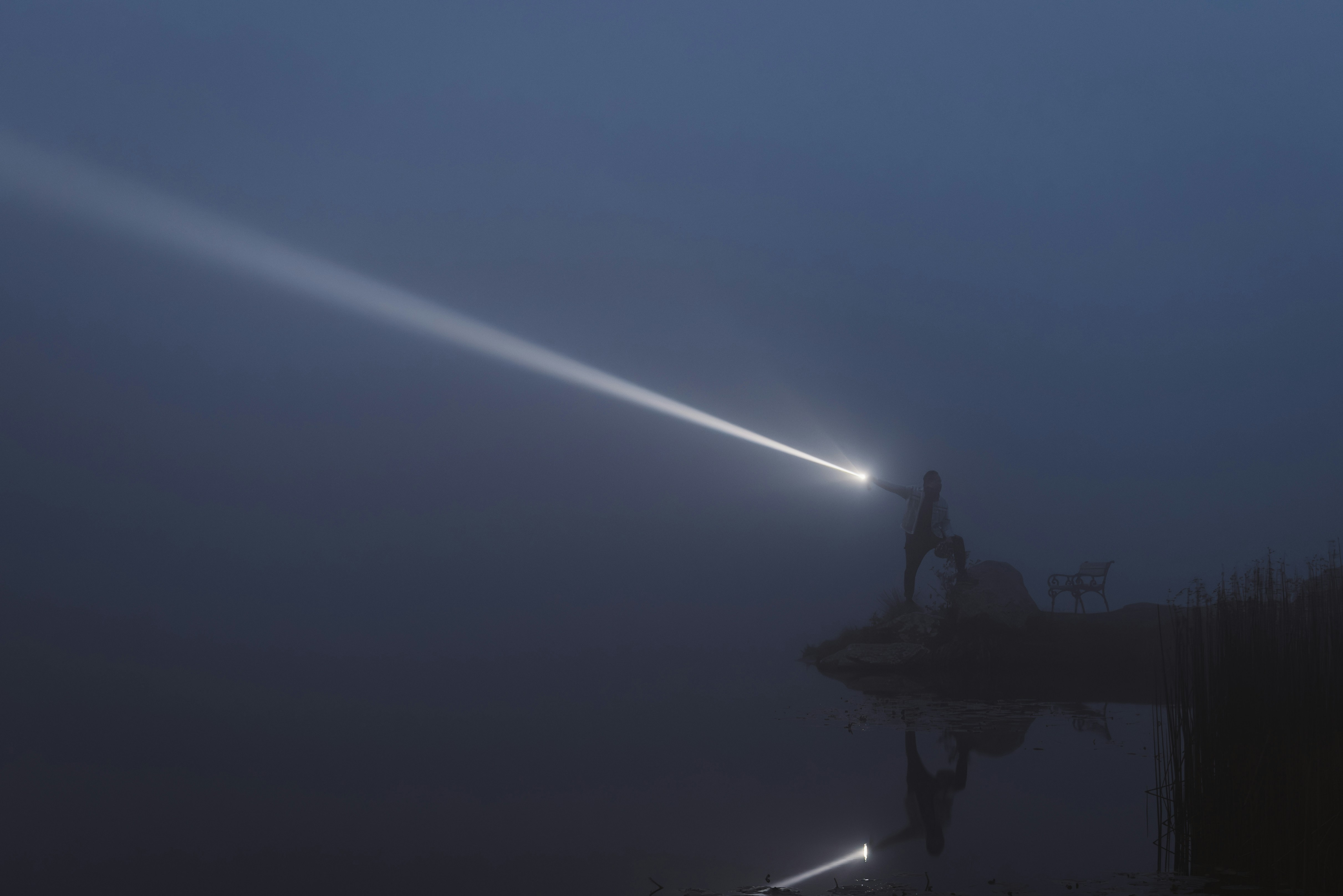 Person holding a flashlight on a foggy lakeside, beam reflecting on the water.