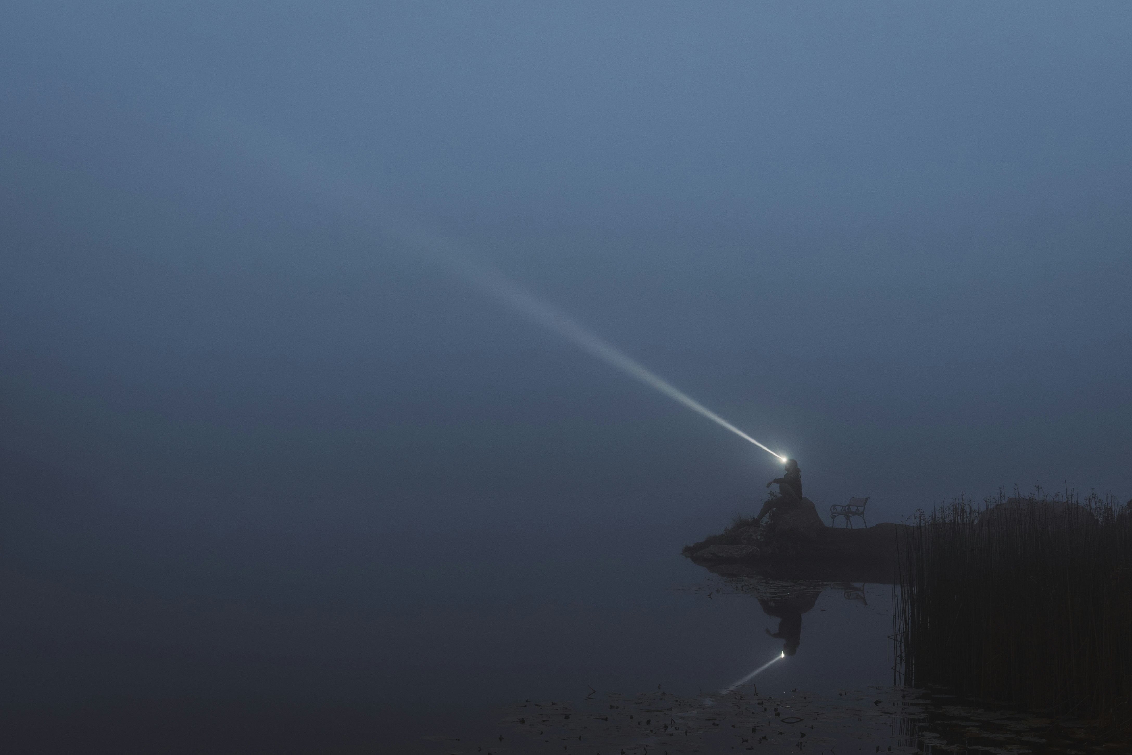 Person with a flashlight on a rock outcrop illuminates a foggy landscape, reflecting in the still water.