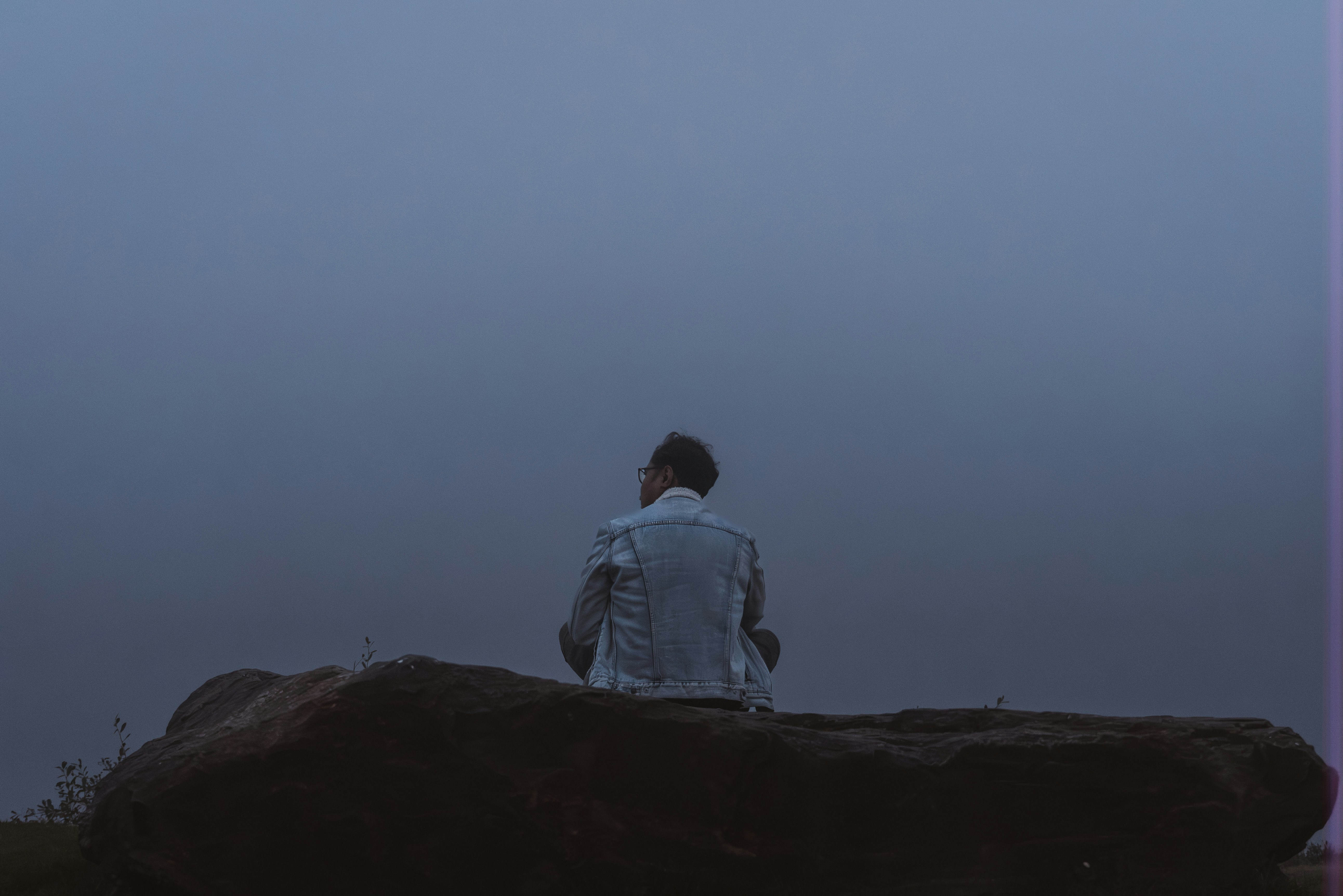 Solitary person in a denim jacket sits on a large rock enveloped by misty blue-gray hues, evoking introspection.
