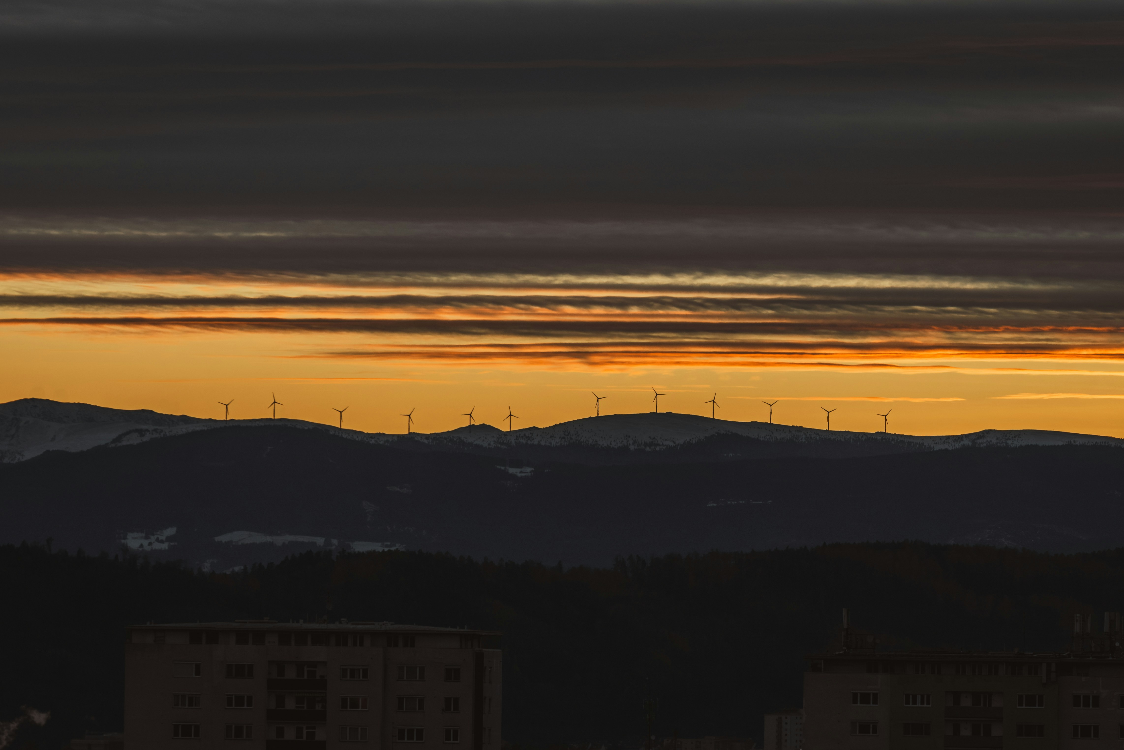 Wind turbines on a distant ridge silhouetted against a layered sunset sky with dark clouds and warm orange hues.