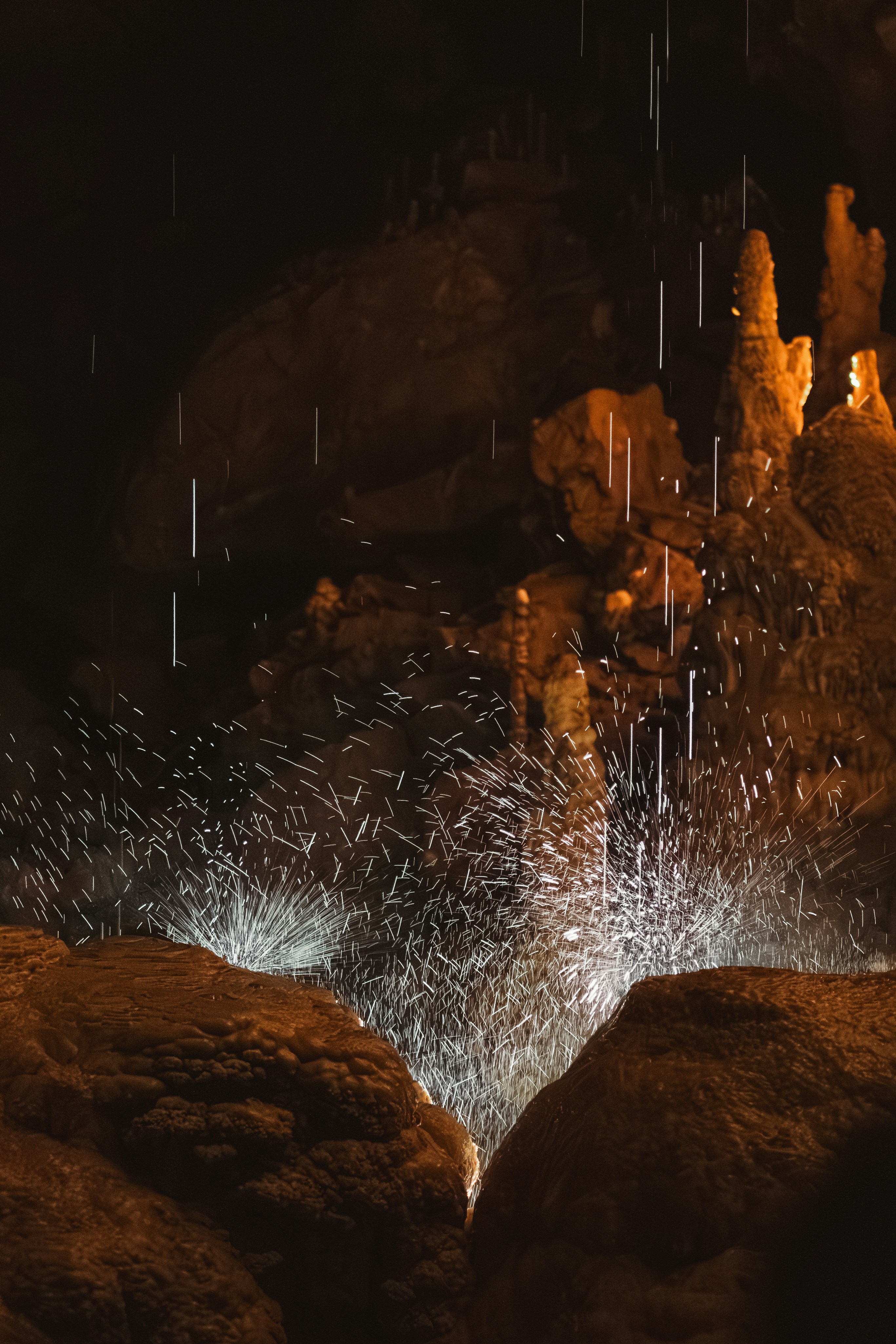 Glowing embers and mineral formations illuminate a dark cave, with sparks arcing upward from a subterranean eruption.