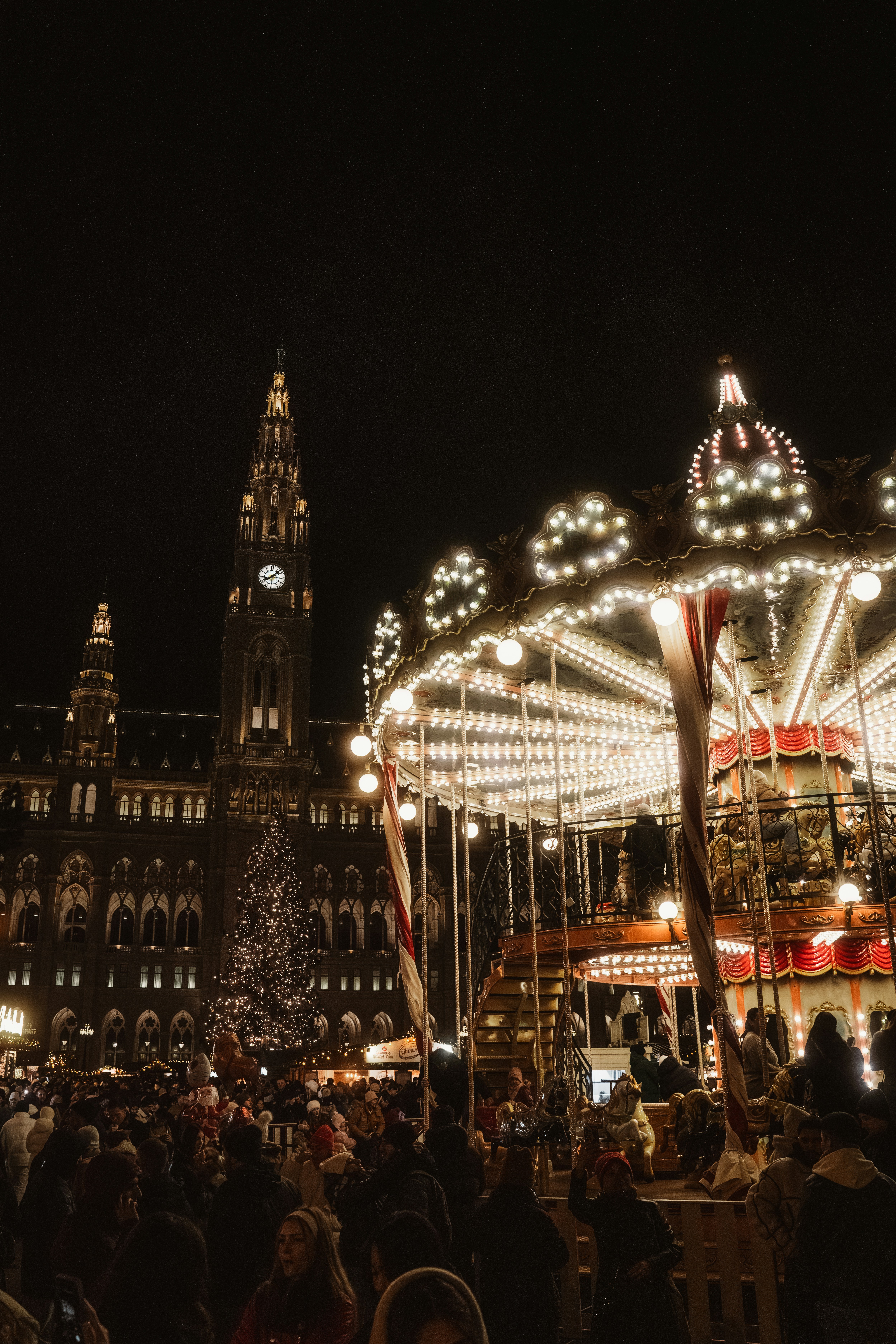 A festive carousel illuminated with bright lights amidst a bustling Christmas market, framed by the majestic architecture of a historic building and a towering Christmas tree.