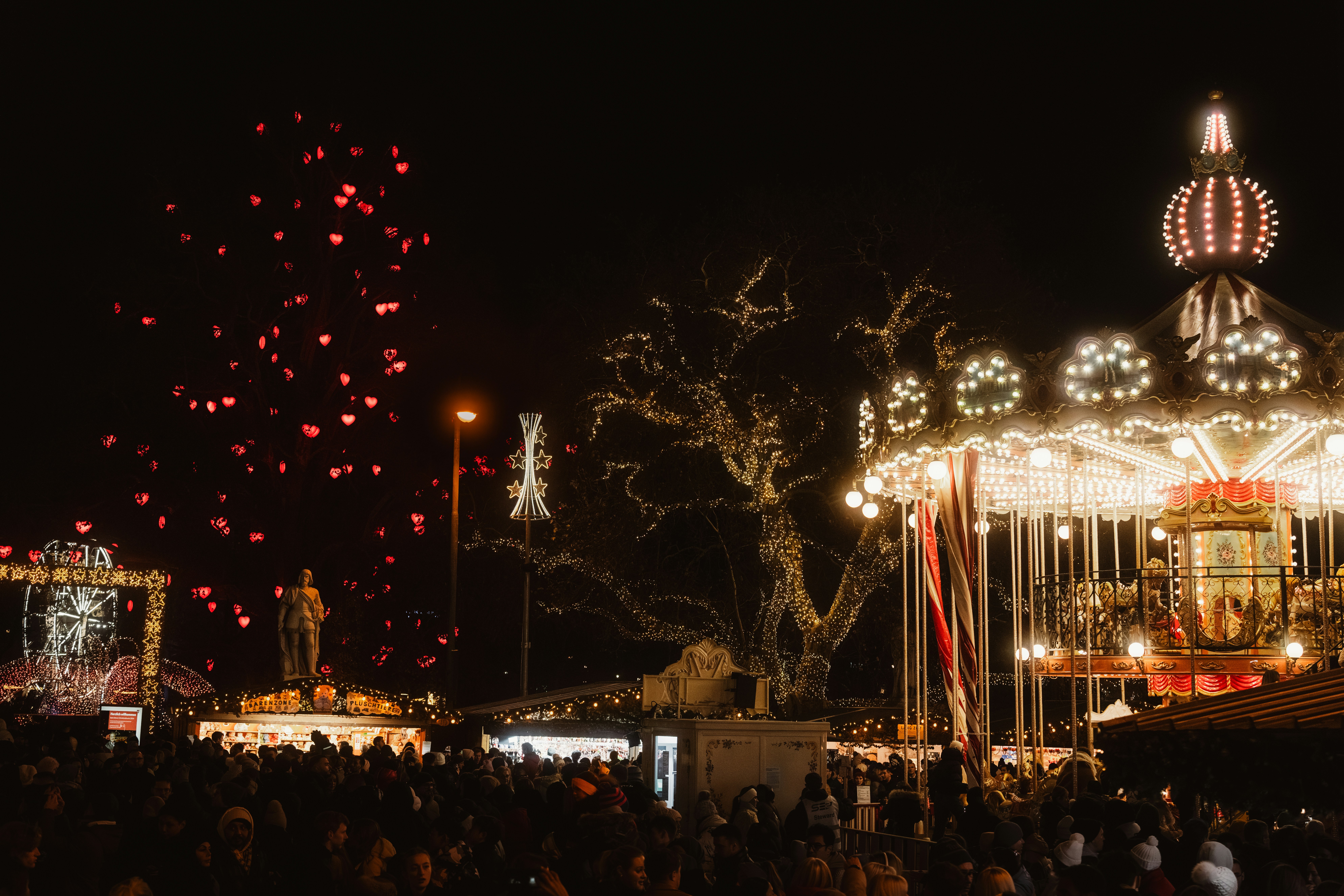 A bustling festive scene featuring a carousel illuminated by lights, surrounded by a crowd and floating heart-shaped lanterns in the night sky.