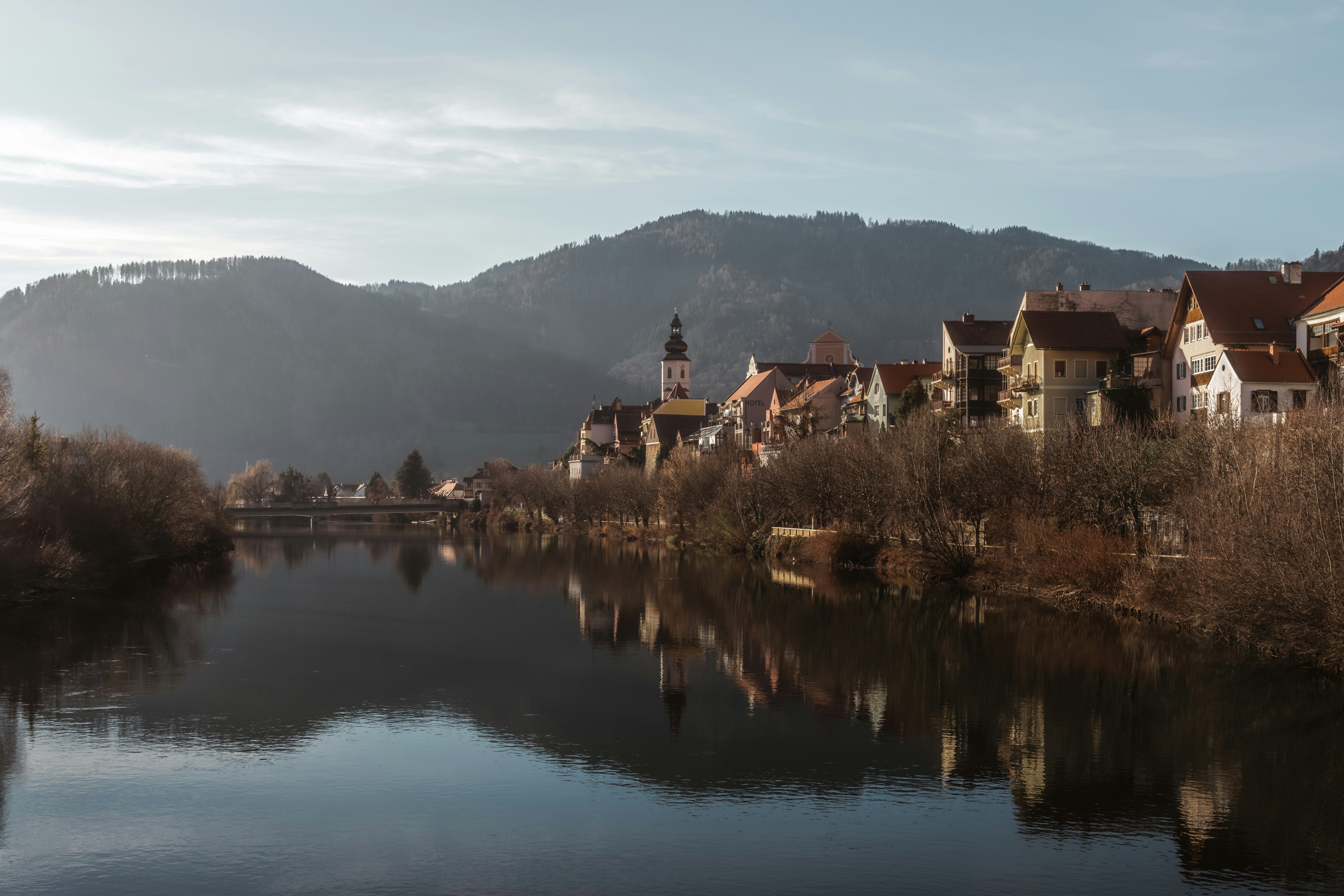Serene village by a calm river with reflections of quaint buildings and bare trees under a soft morning light.