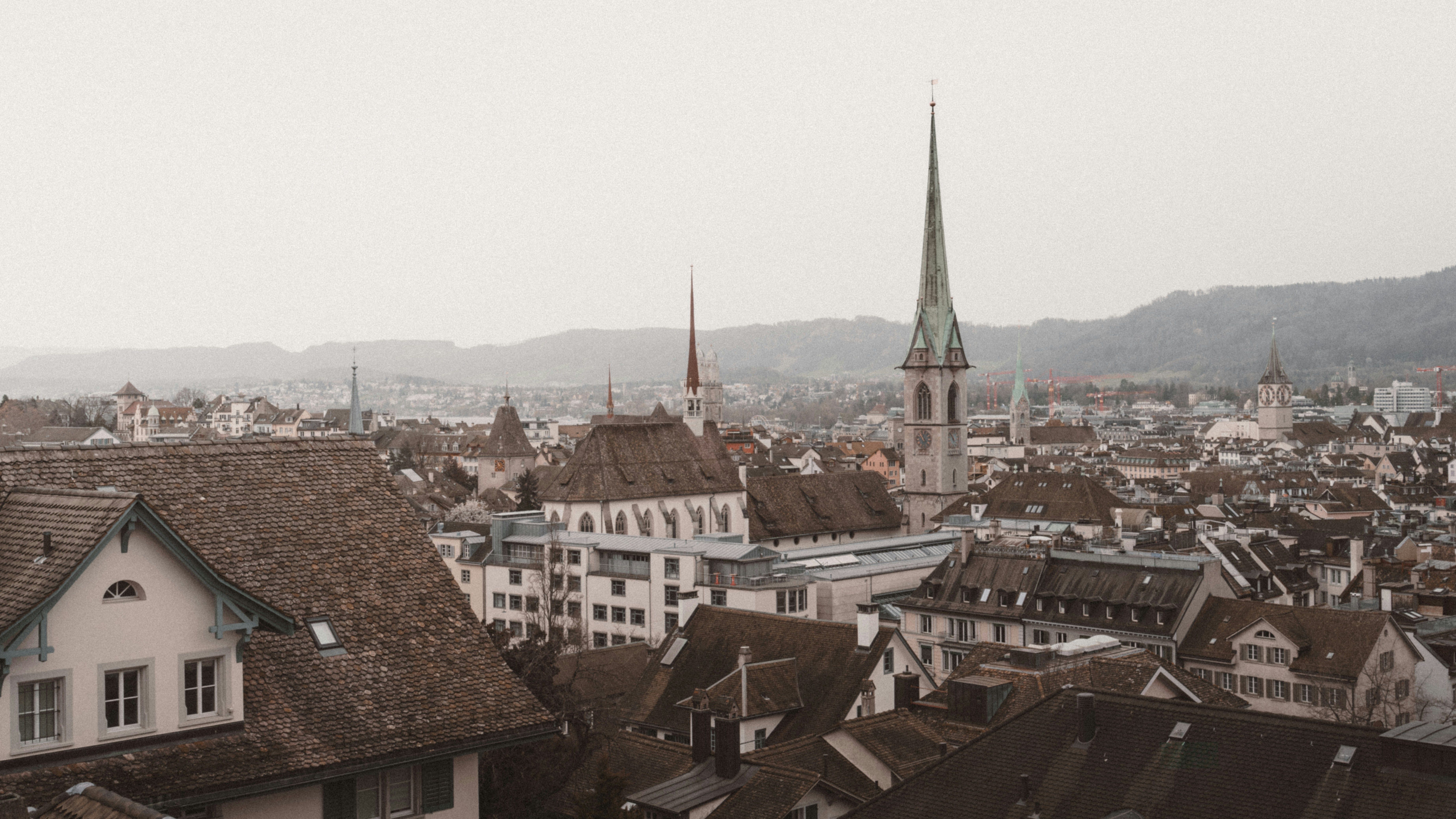 Historic city skyline with church spires rising above rustic rooftops against a muted, overcast sky.