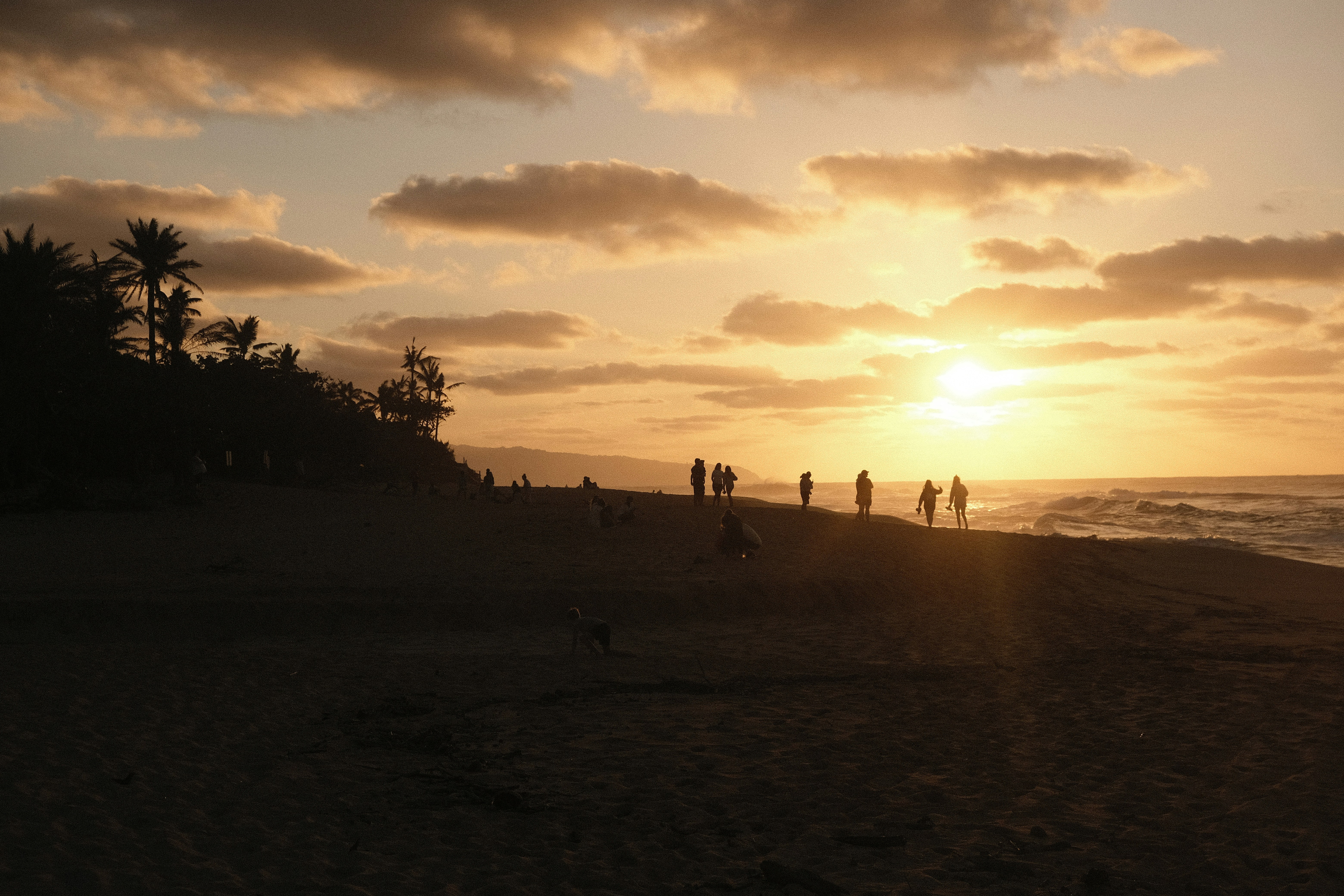 Silhouetted figures walk along a beach during a vibrant sunset with scattered clouds.