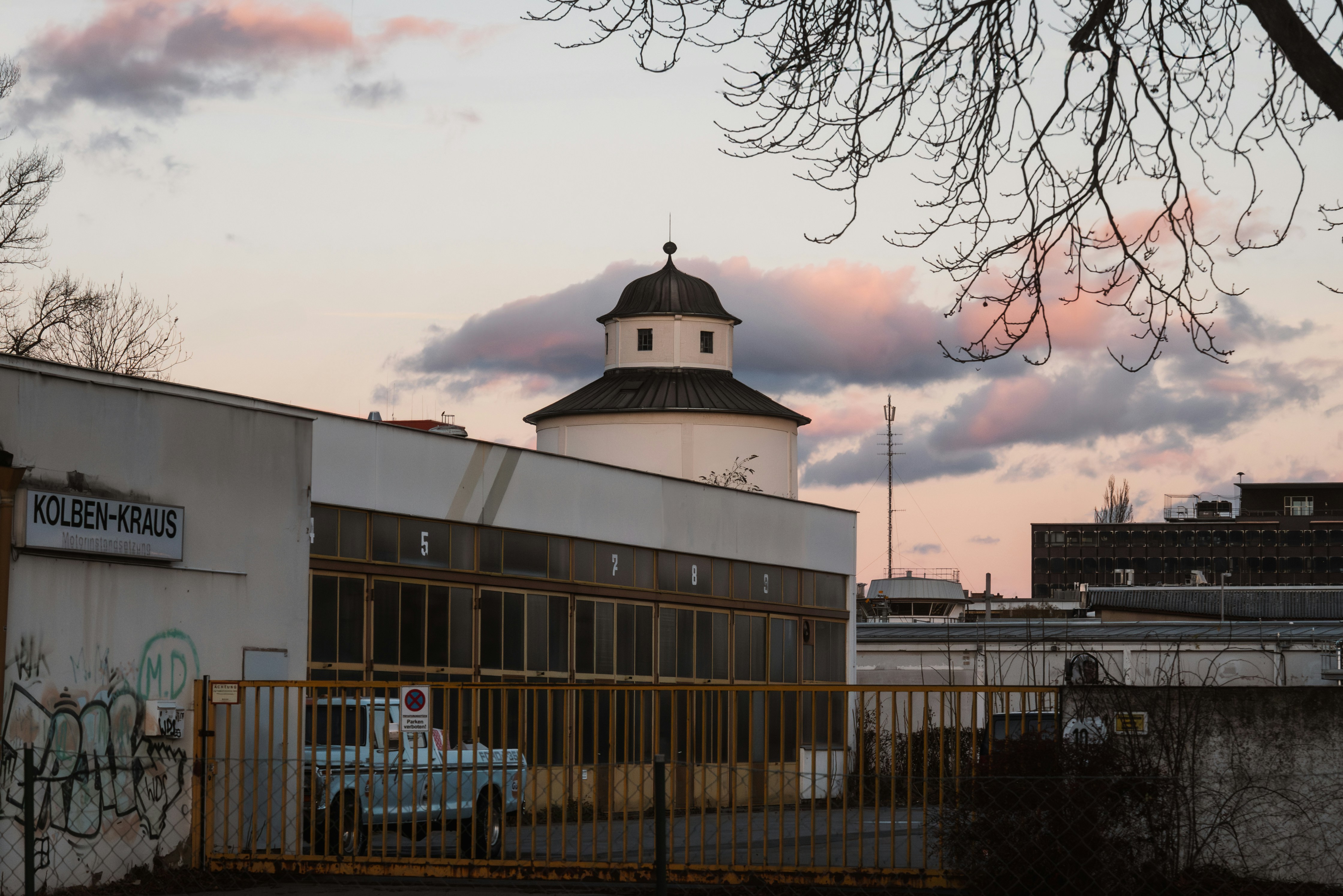 Historic tower rises above industrial buildings under a pastel sunset sky, framed by tree branches.