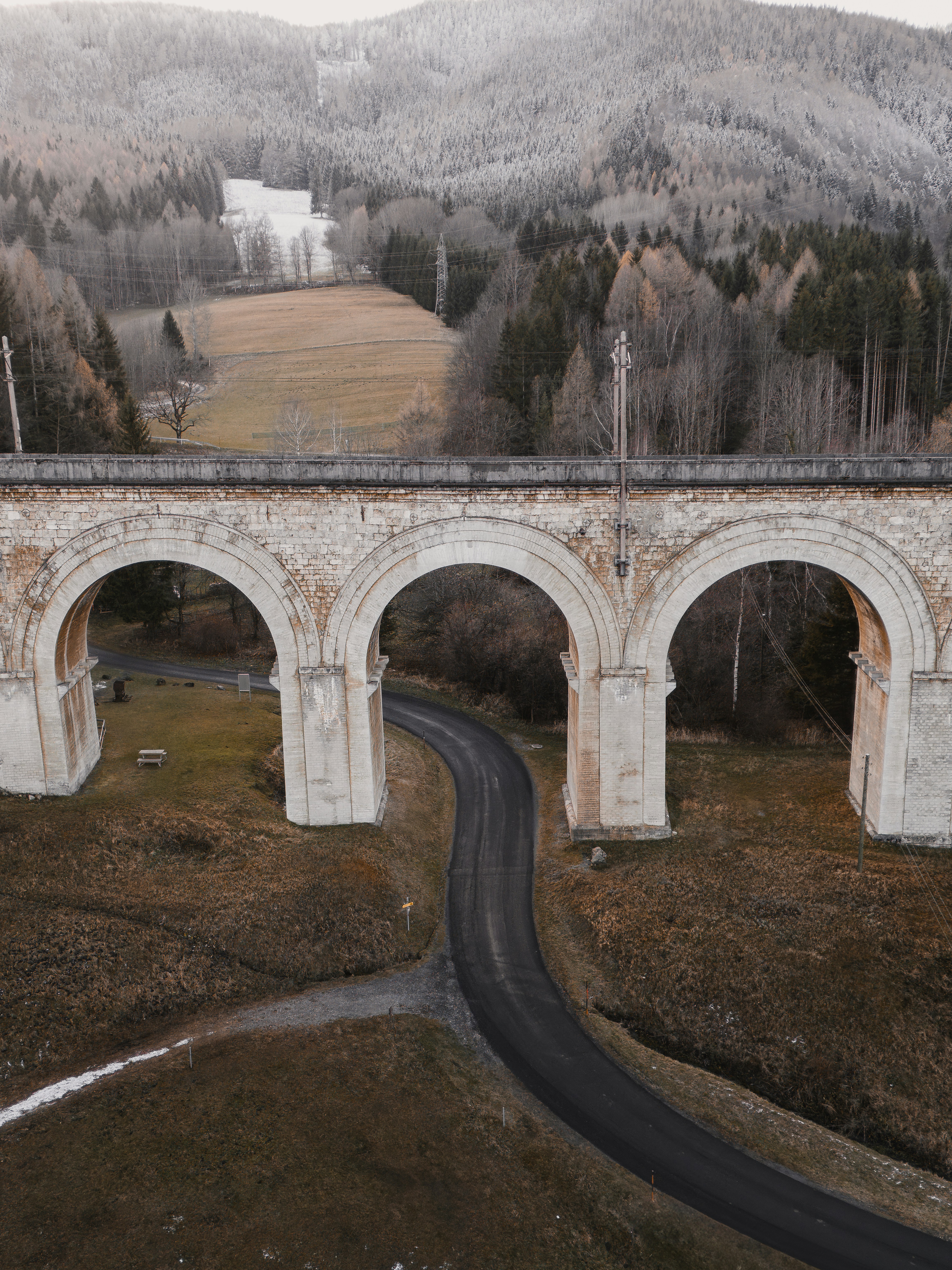 Aerial shot of a stone viaduct with three large arches spanning a winding road through a valley, framed by forested hills and distant fields.