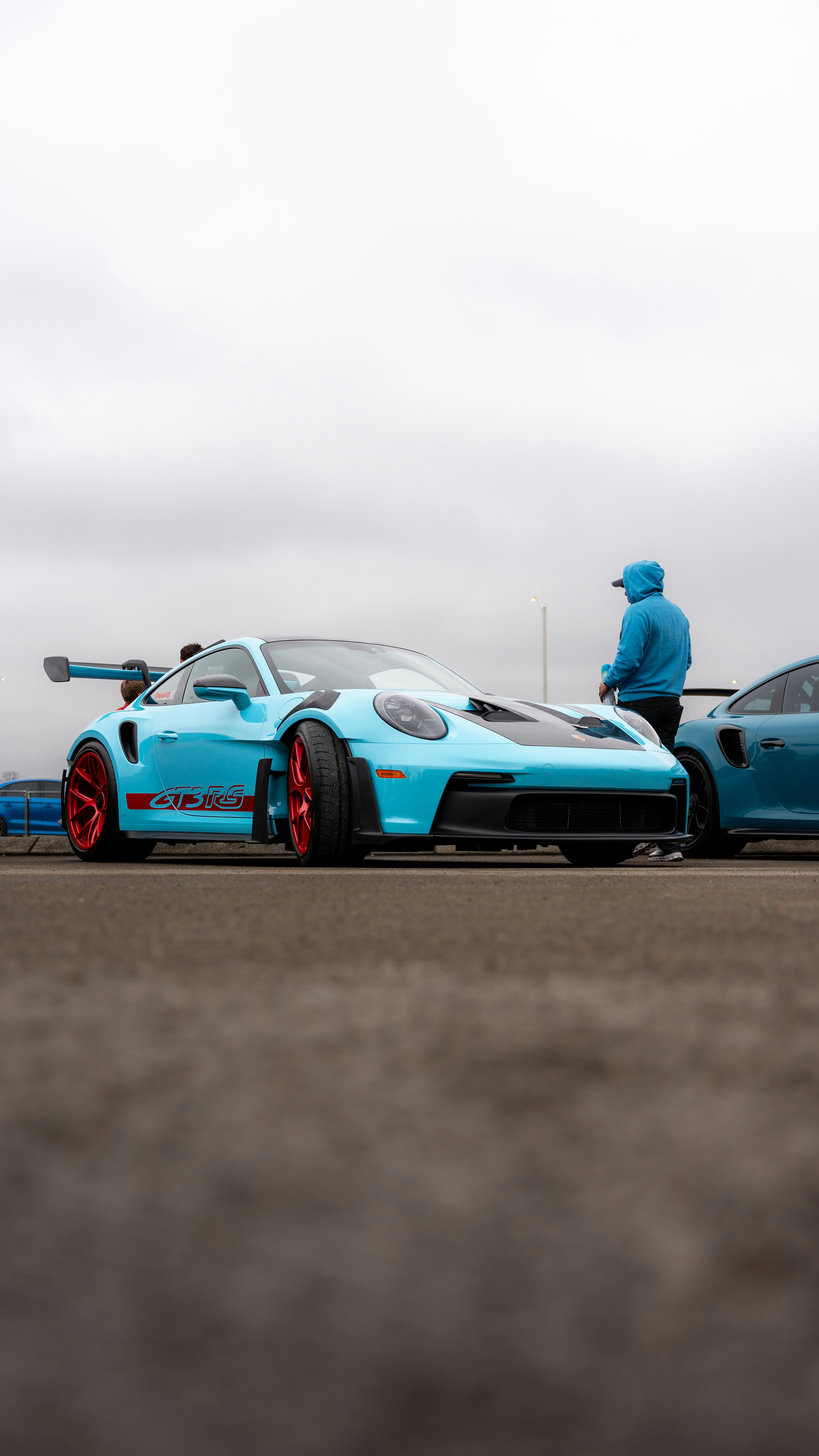 A rare blue and red GT3 RS with its driver walking along the far side wearing a similar blue hoodie. The sky is grey and dull, allowing for the car to pop out from the background and foreground.