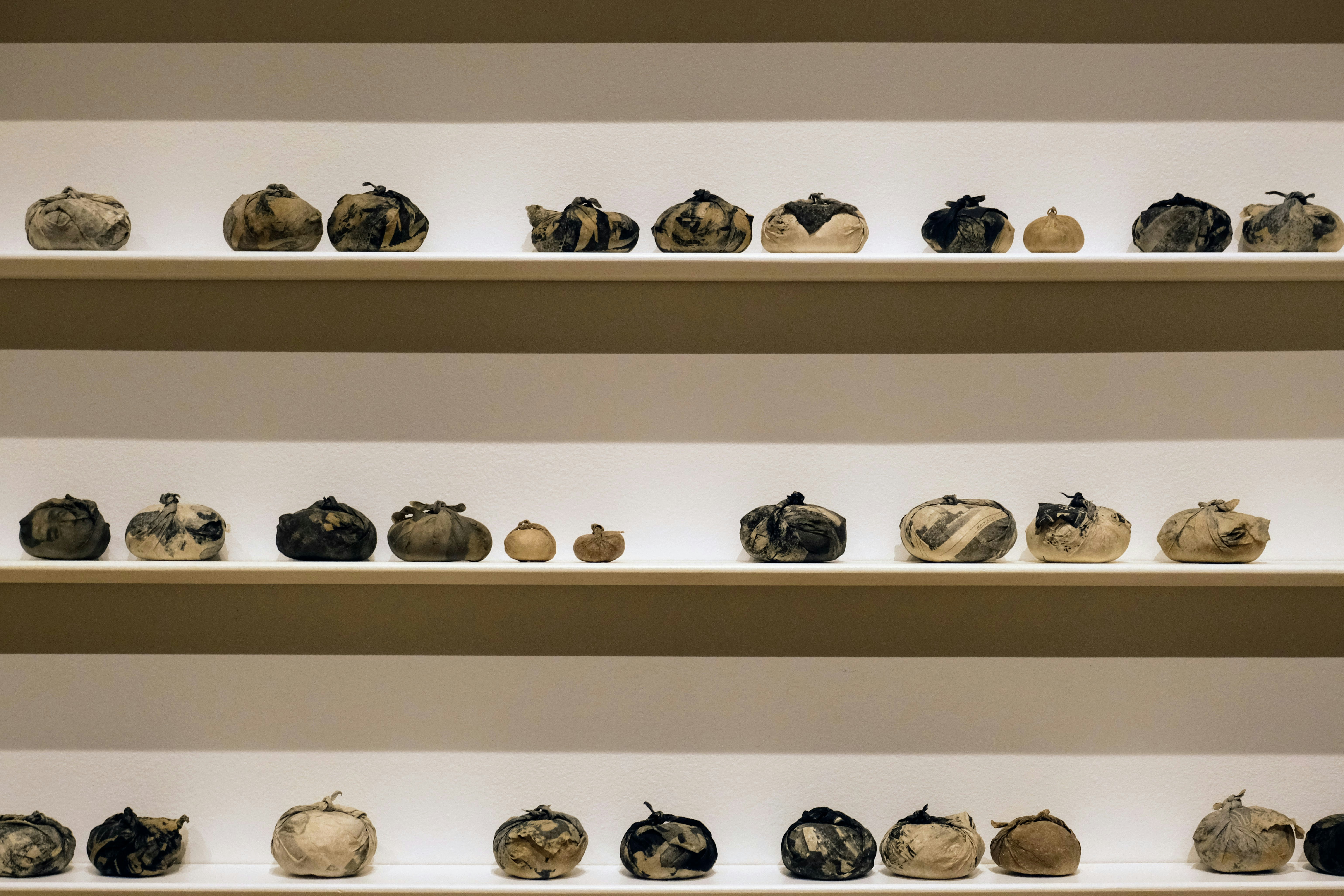 Uniquely wrapped bundles in beige and black rest on minimalist white shelves, creating a balanced composition with soft, diffused lighting.